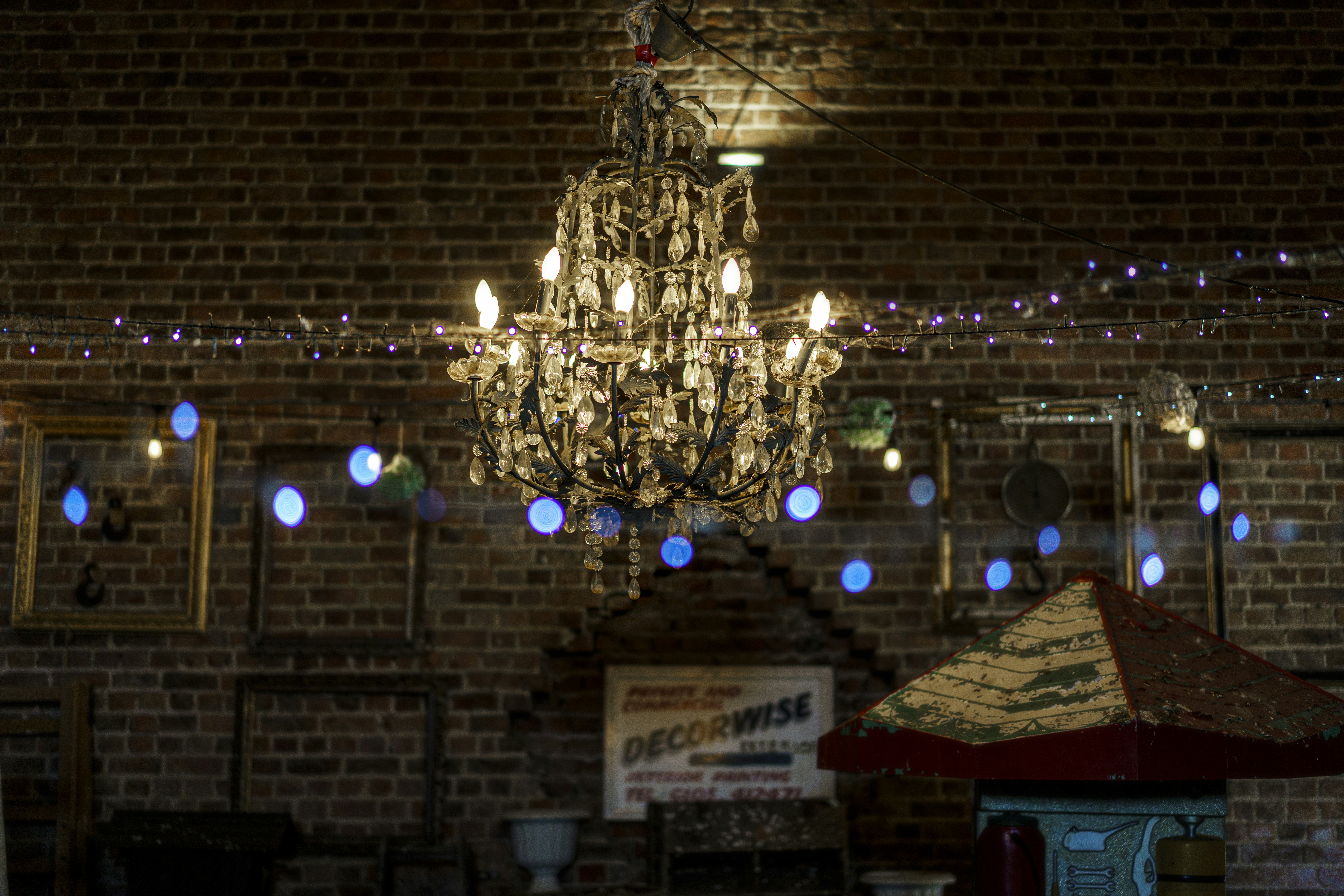 A crystal chandelier hanging over a brick-walled interior with string lights and ceiling decor, creating a warm, festive ambiance.