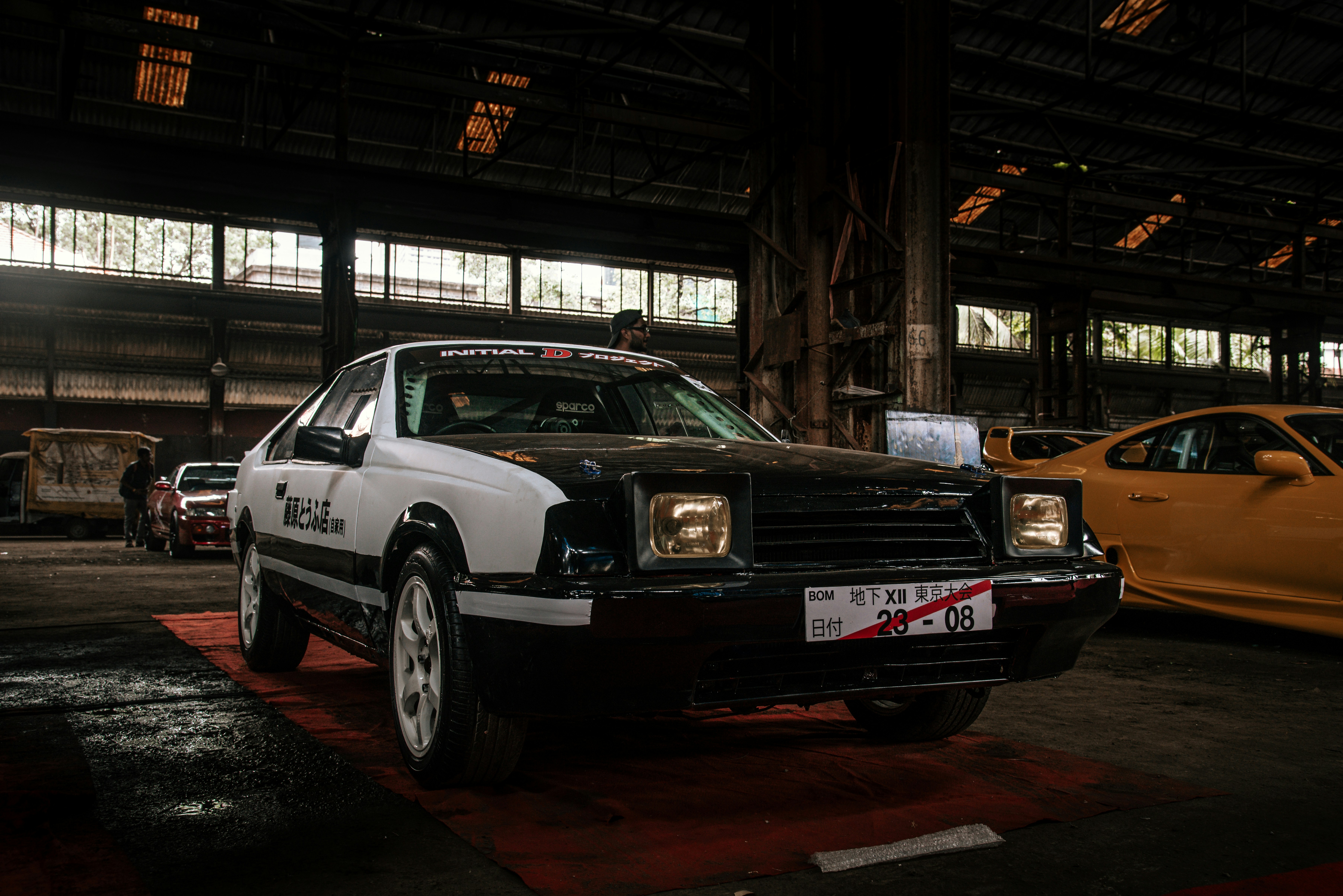 White vintage racing taxi sits on a red mat inside a dim warehouse, with other colorful cars in the background.