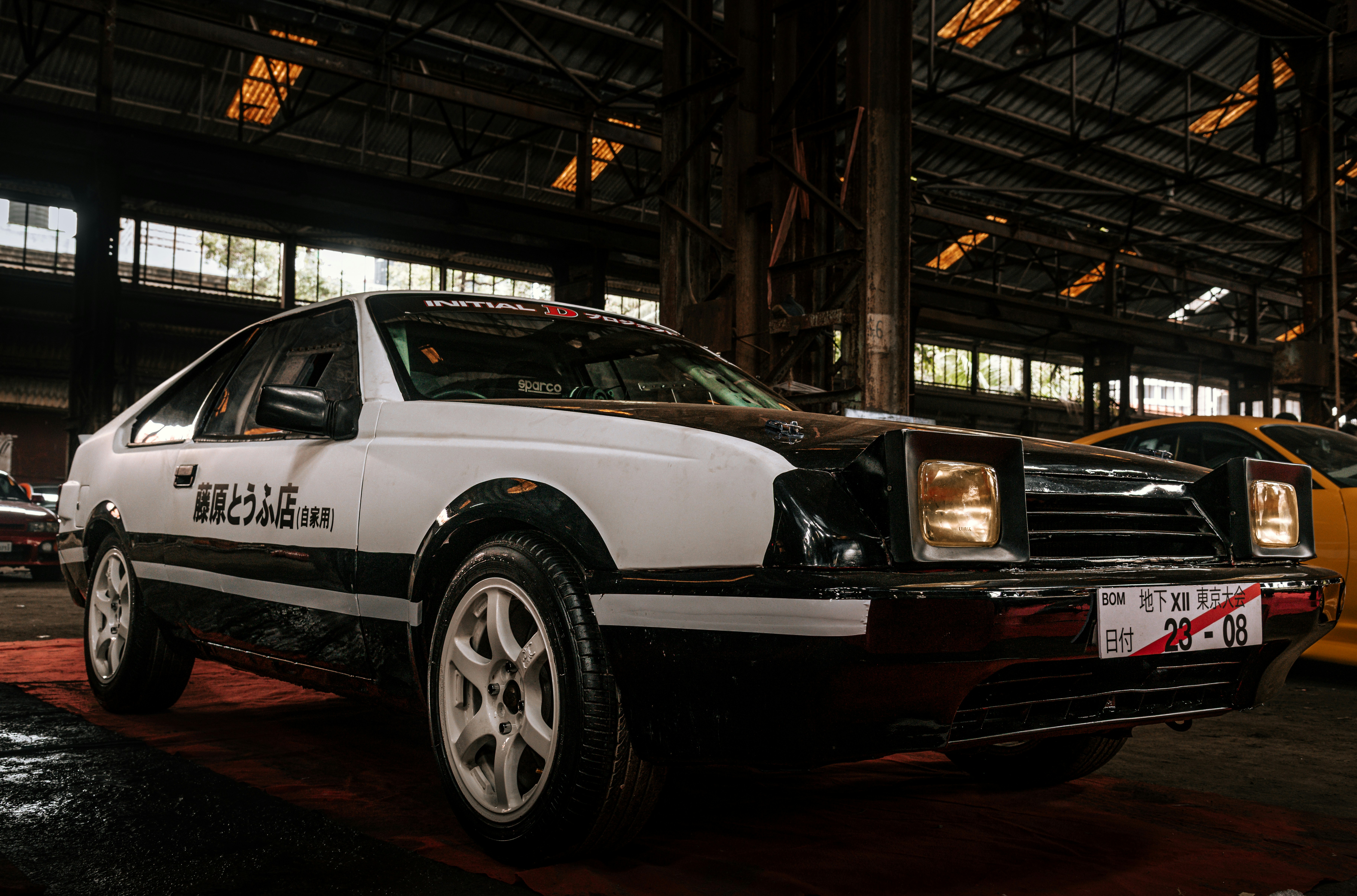 A white vintage sedan with black trim sits in a dark industrial space, showing Japanese characters along the side; the front grille and headlights are prominent, suggesting a workshop or showroom setting.