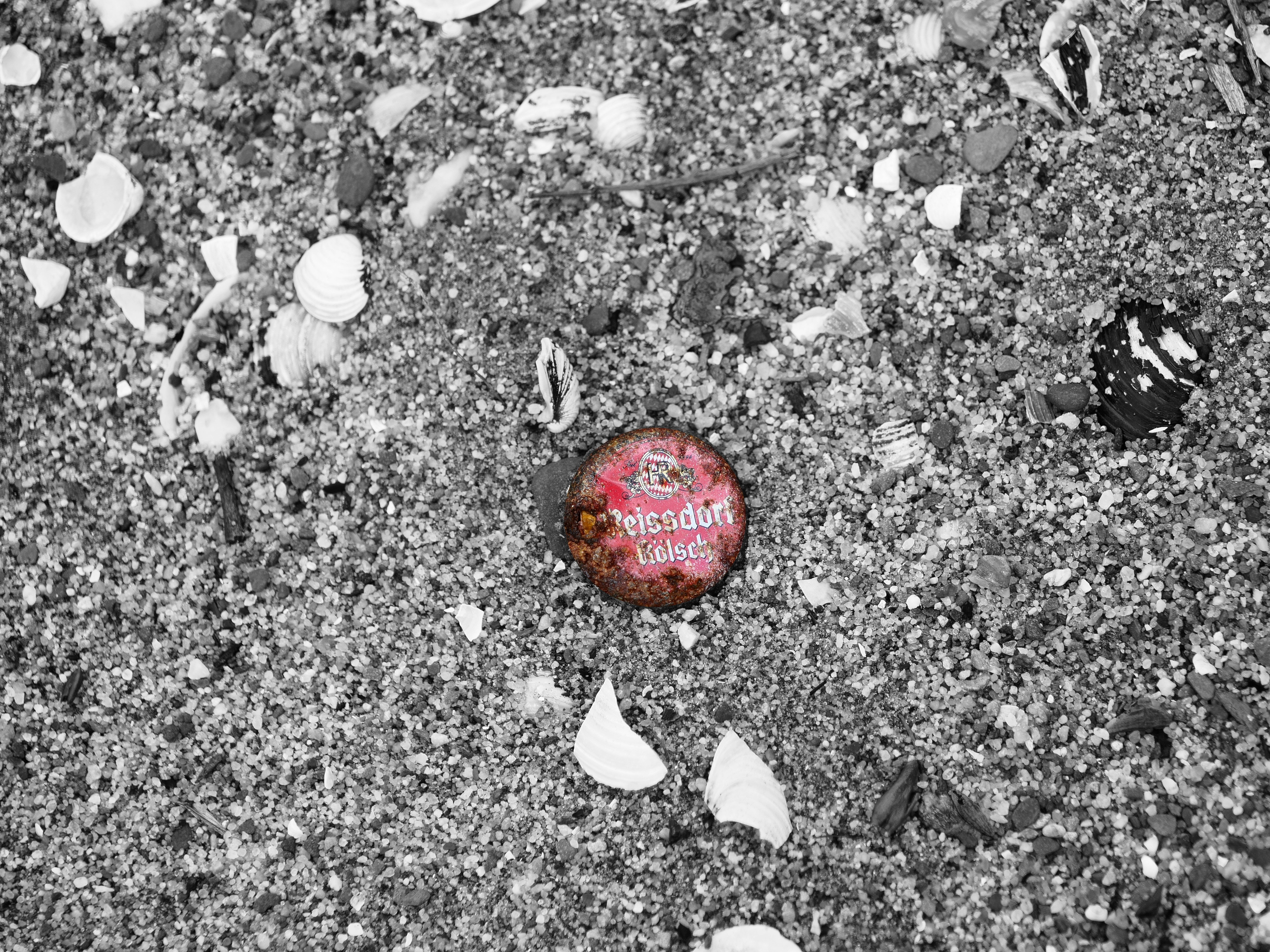 Close-up of a red bottle cap lying in gritty sand with small shells and specks; the cap appears weathered and worn, drawing attention as the focal point of the ground texture. Logo-like object in a monochrome foreground.