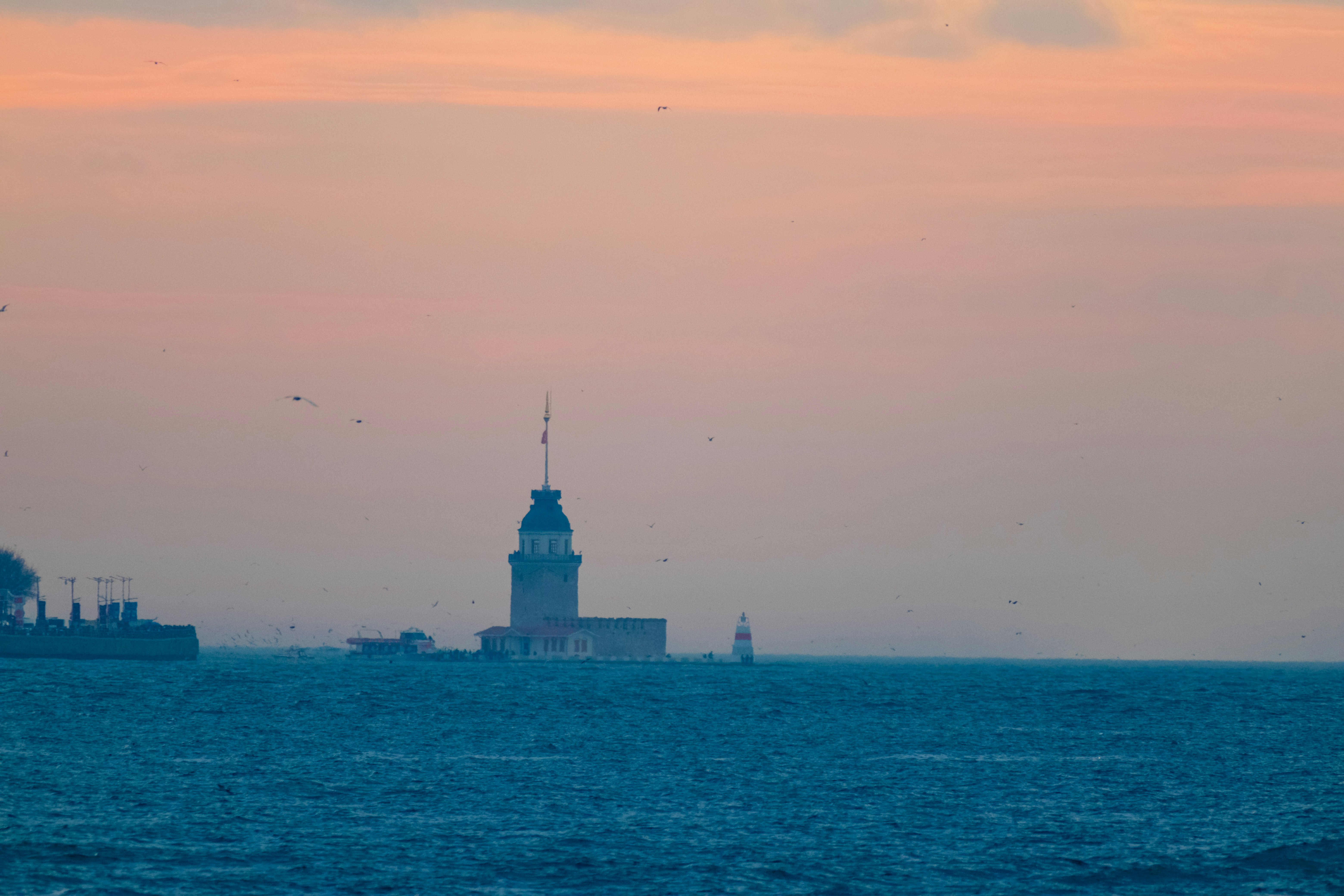 Evening light washes the horizon as a solitary lighthouse rises from calm sea, silhouetted against a pastel sky.