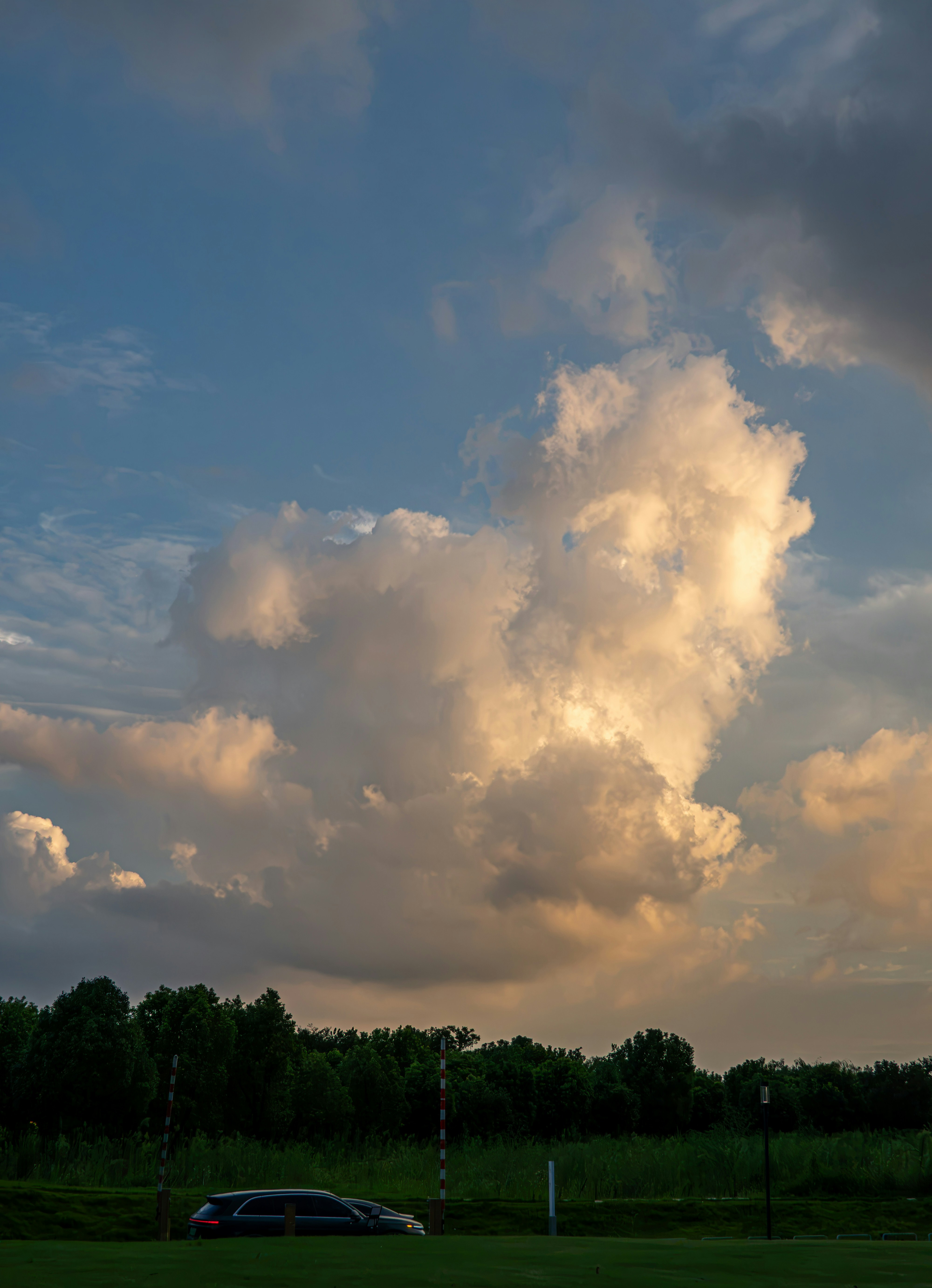 Golden-illuminated cumulus cloud towering above a flat, grassy field with distant trees and a parked car, emphasizing vast sky and calm landscape.