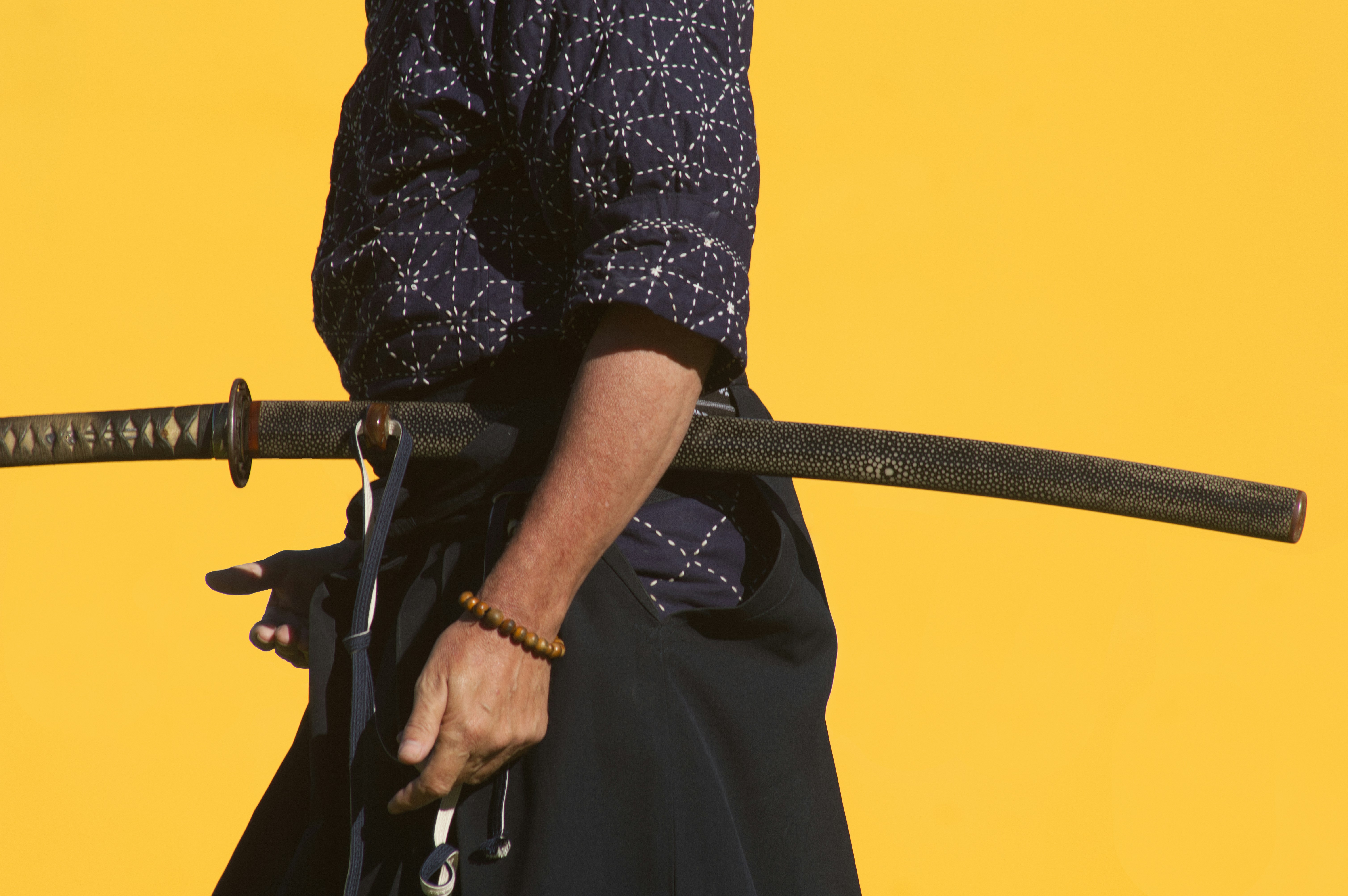 A close crop of a person holding a sheathed sword against a bold yellow background, focusing on the torso and hands with the blade resting across the midsection.