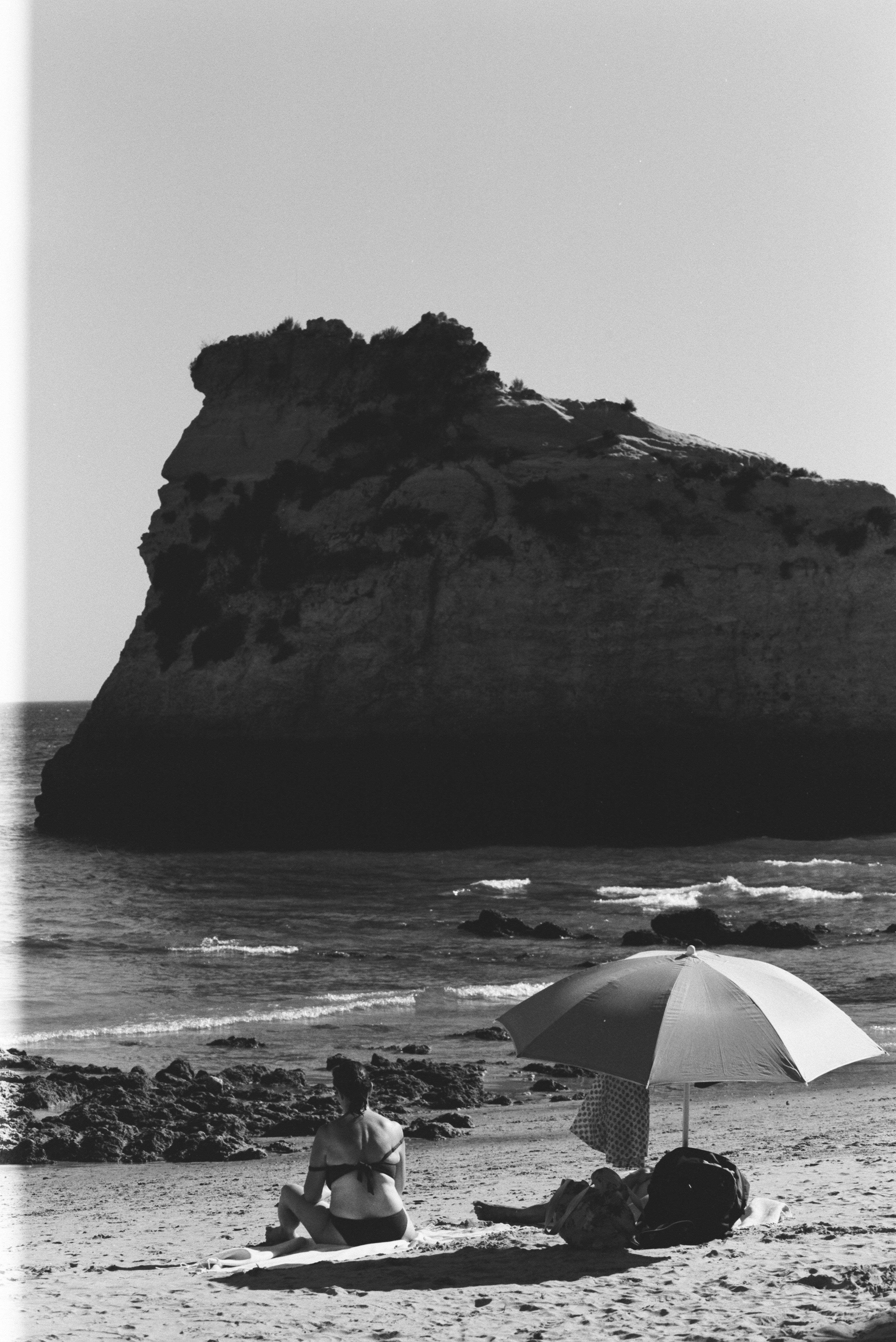 Monochrome beach scene with a prominent rock formation in the background and two sunbathers under a large umbrella near the shore.