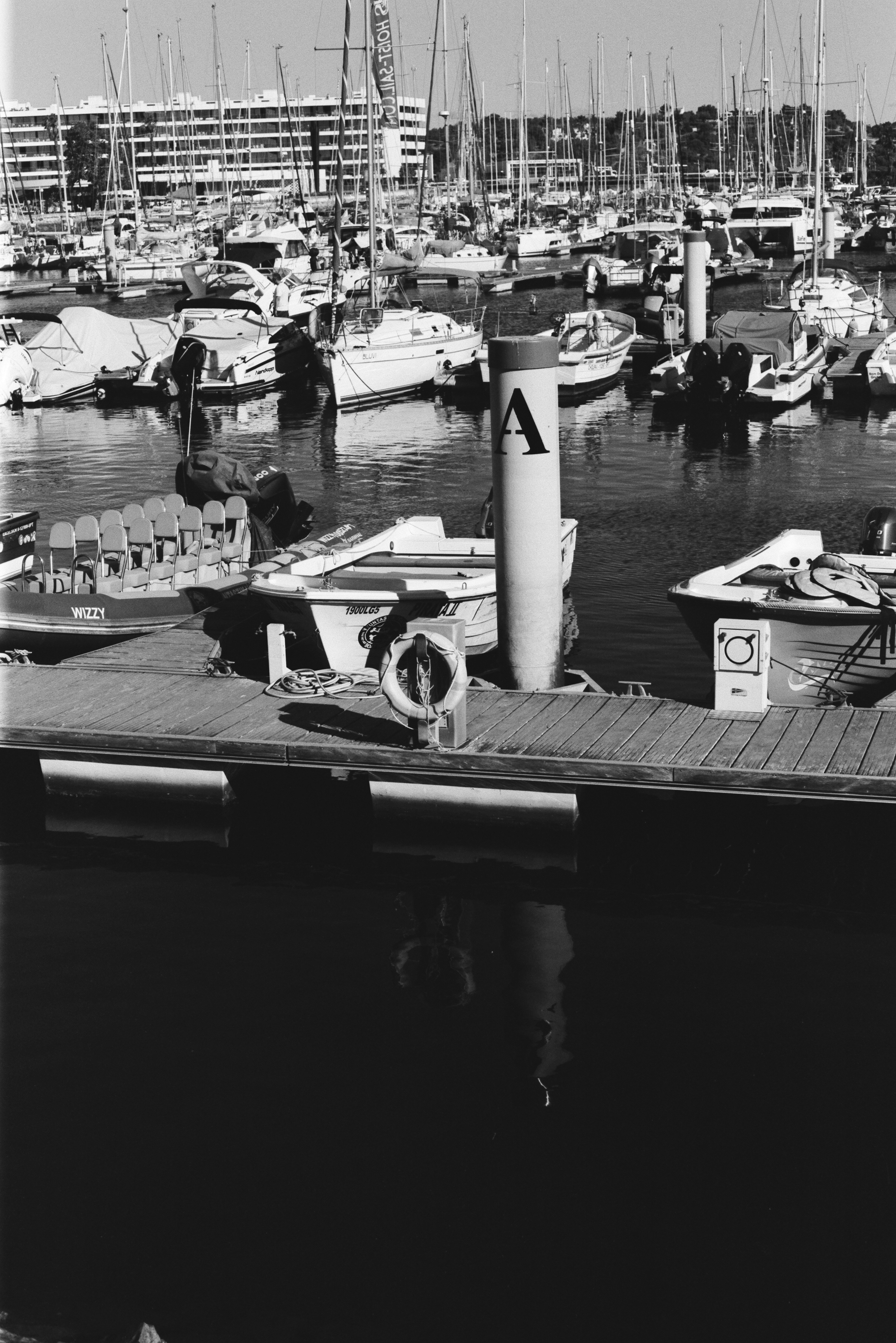 Monochrome marina scene with a wooden dock, moored boats, and a tall A-mark post at the center foreground; the composition emphasizes the dense fleet and reflections on calm water.