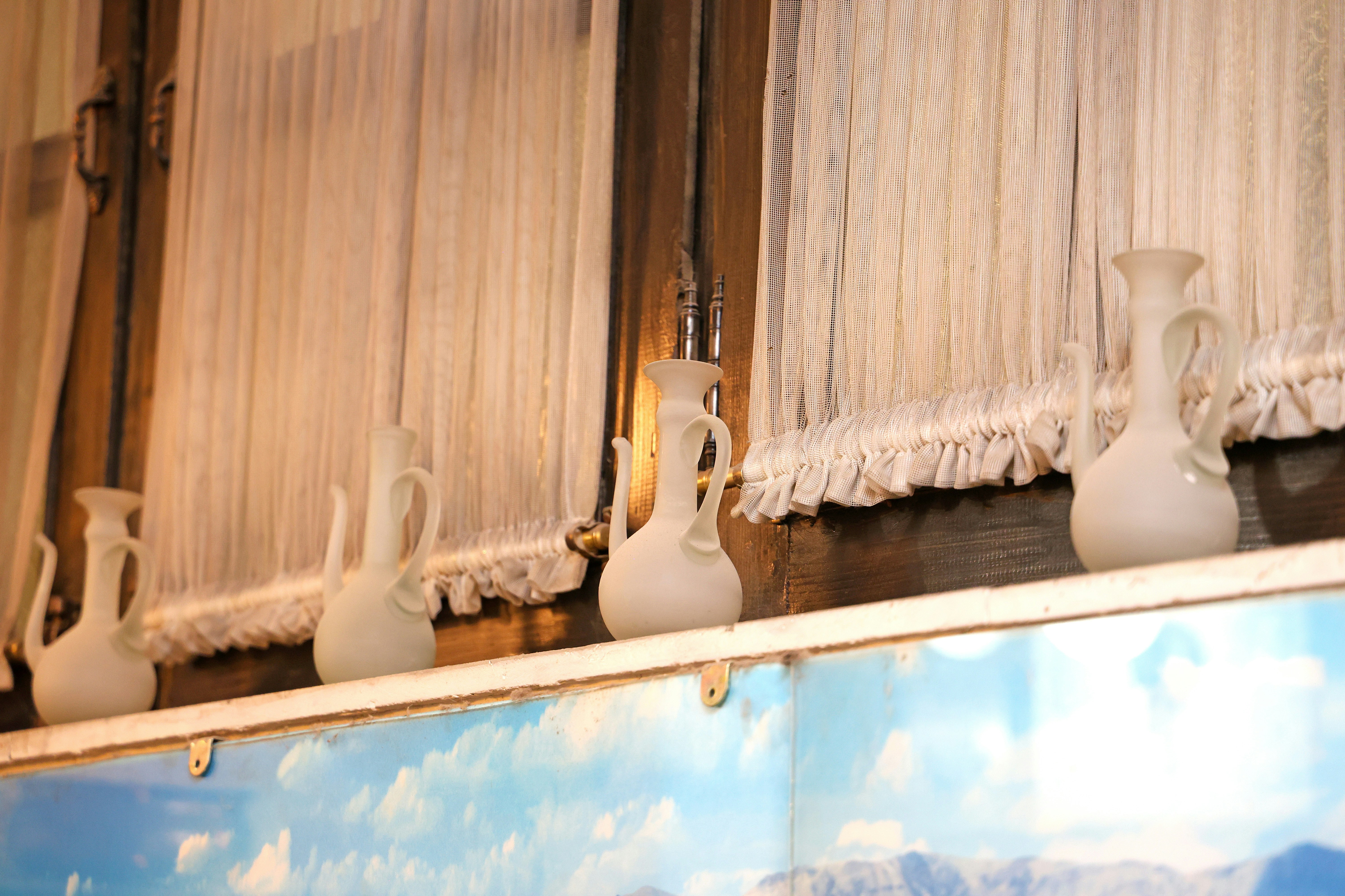Row of small white ceramic flasks lined up on a wooden shelf above a blue sky mural; soft warm ambient lighting and draped curtains in the background.