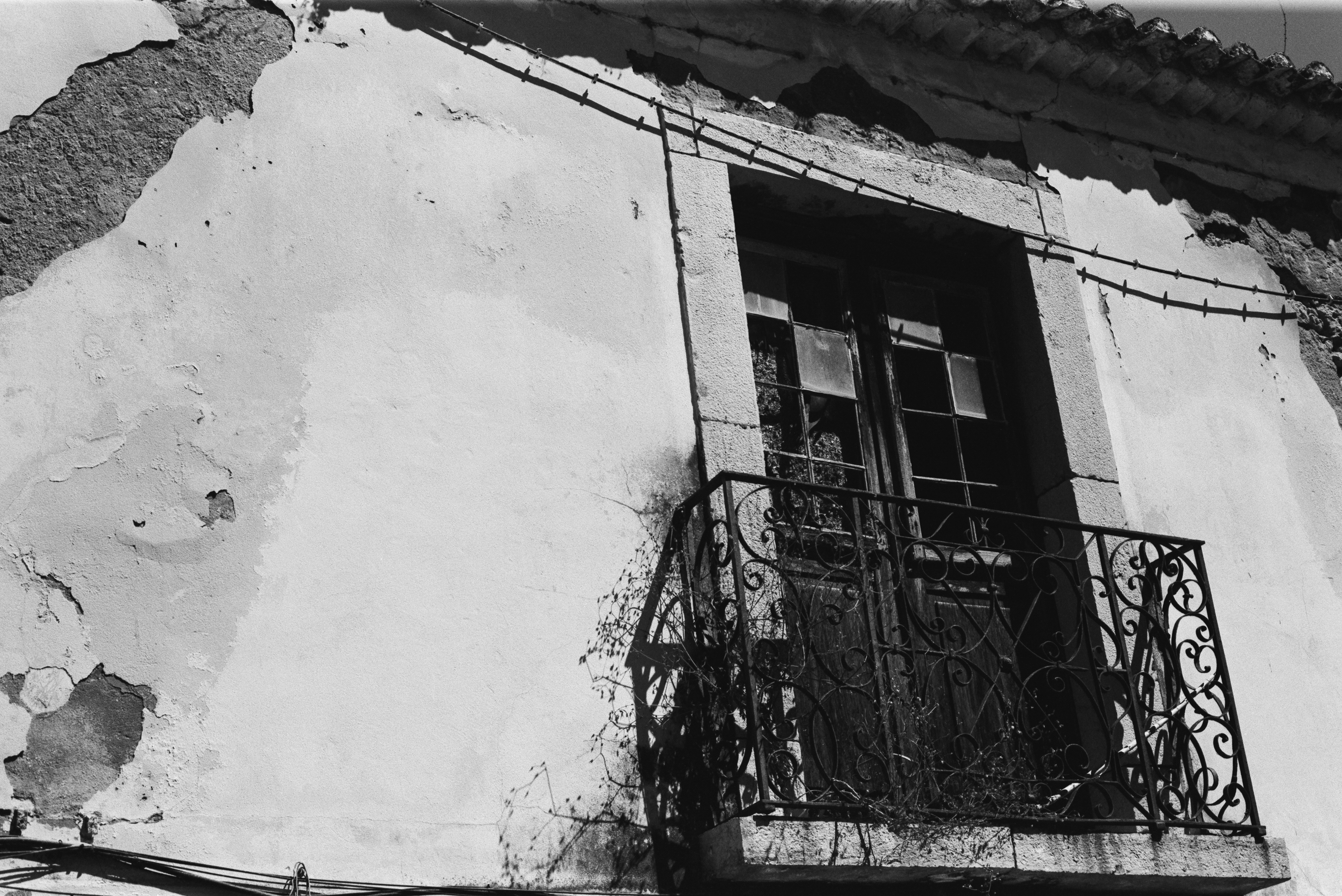 Monochrome exterior of a weathered building featuring a small ornate balcony and a dark door/window, conveying aged urban texture and quiet decay.