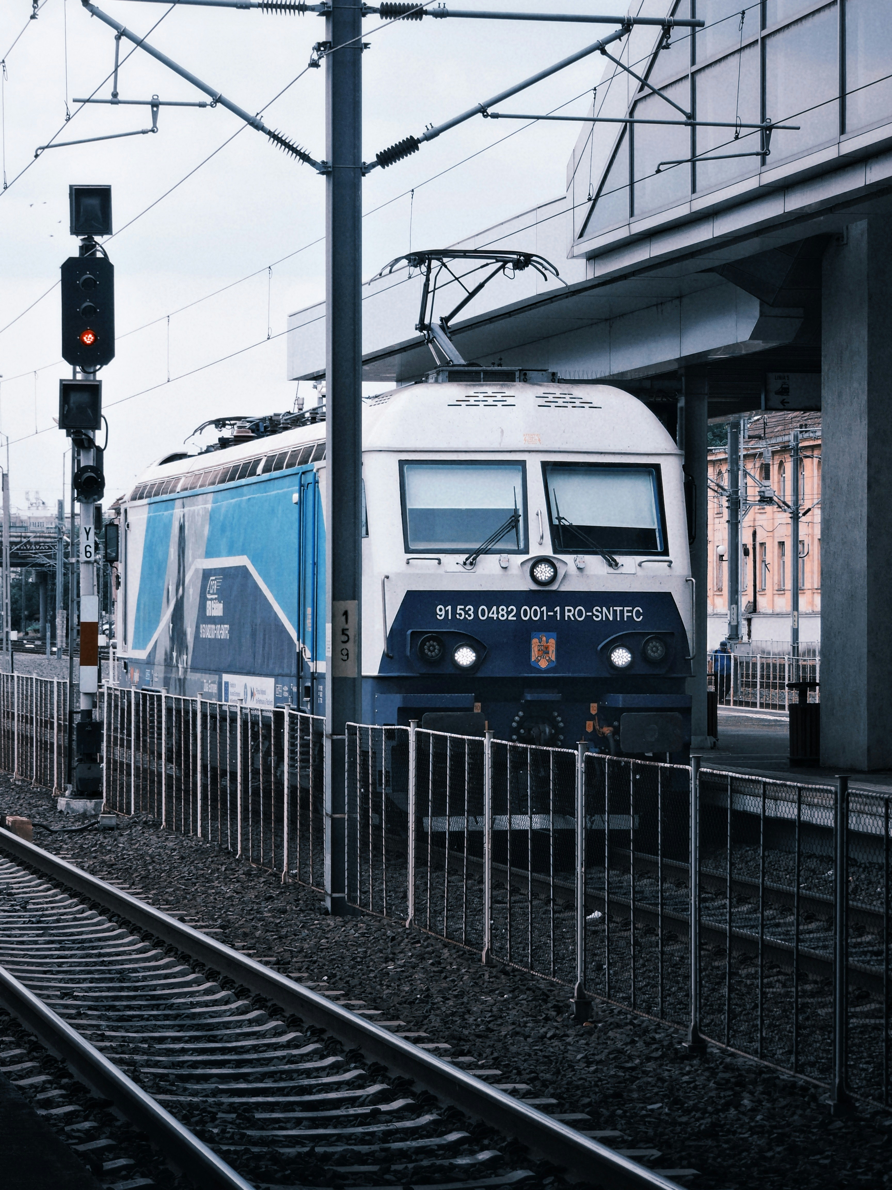 Blue-and-white locomotive sits under a concrete overpass at a quiet station, rails stretching toward the foreground with fencing and signals framing the scene.