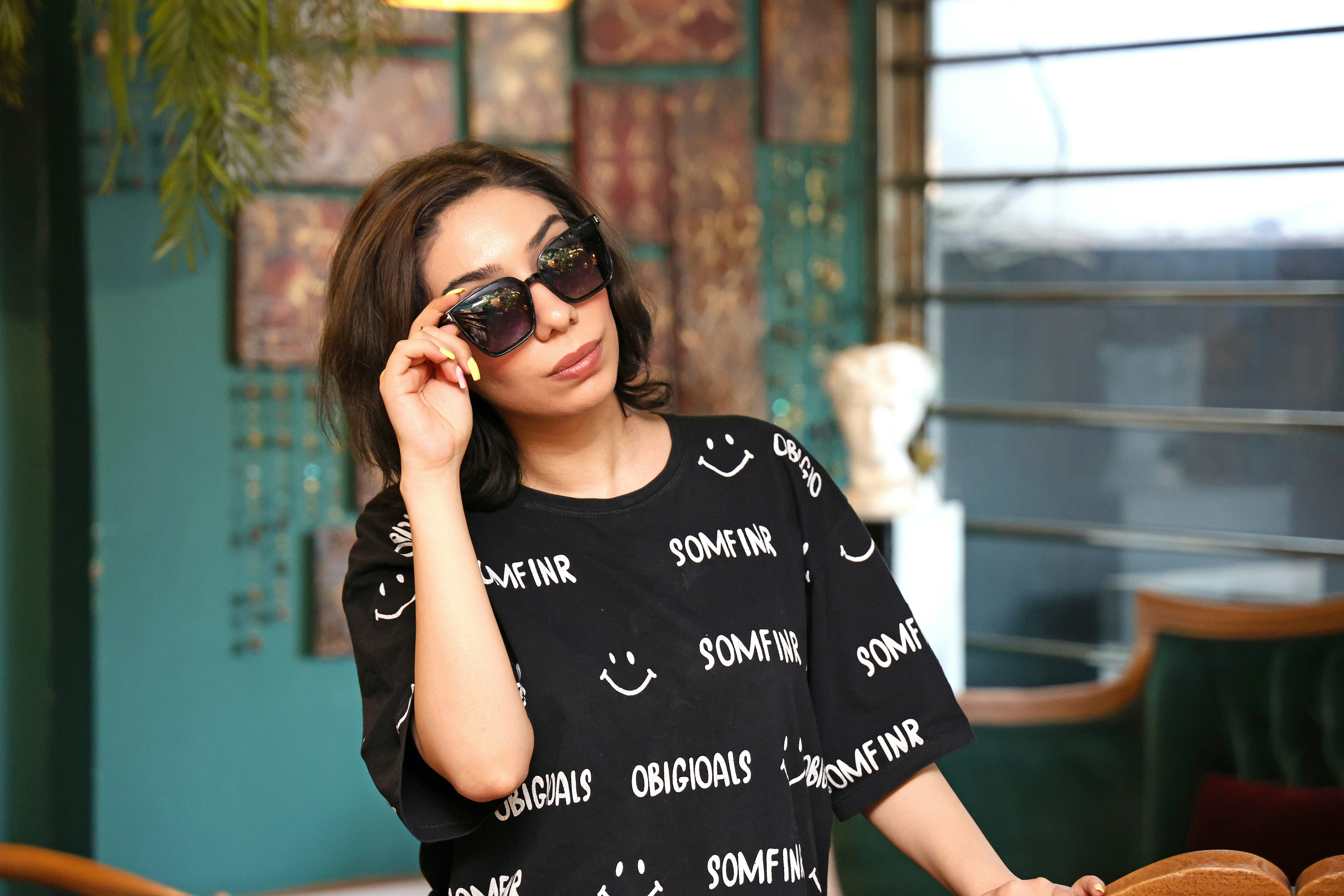 Woman wearing a printed shirt and sunglasses posing casually in a cafe setting. The scene emphasizes her relaxed expression and fashion choice with a colorful, slightly blurred background.