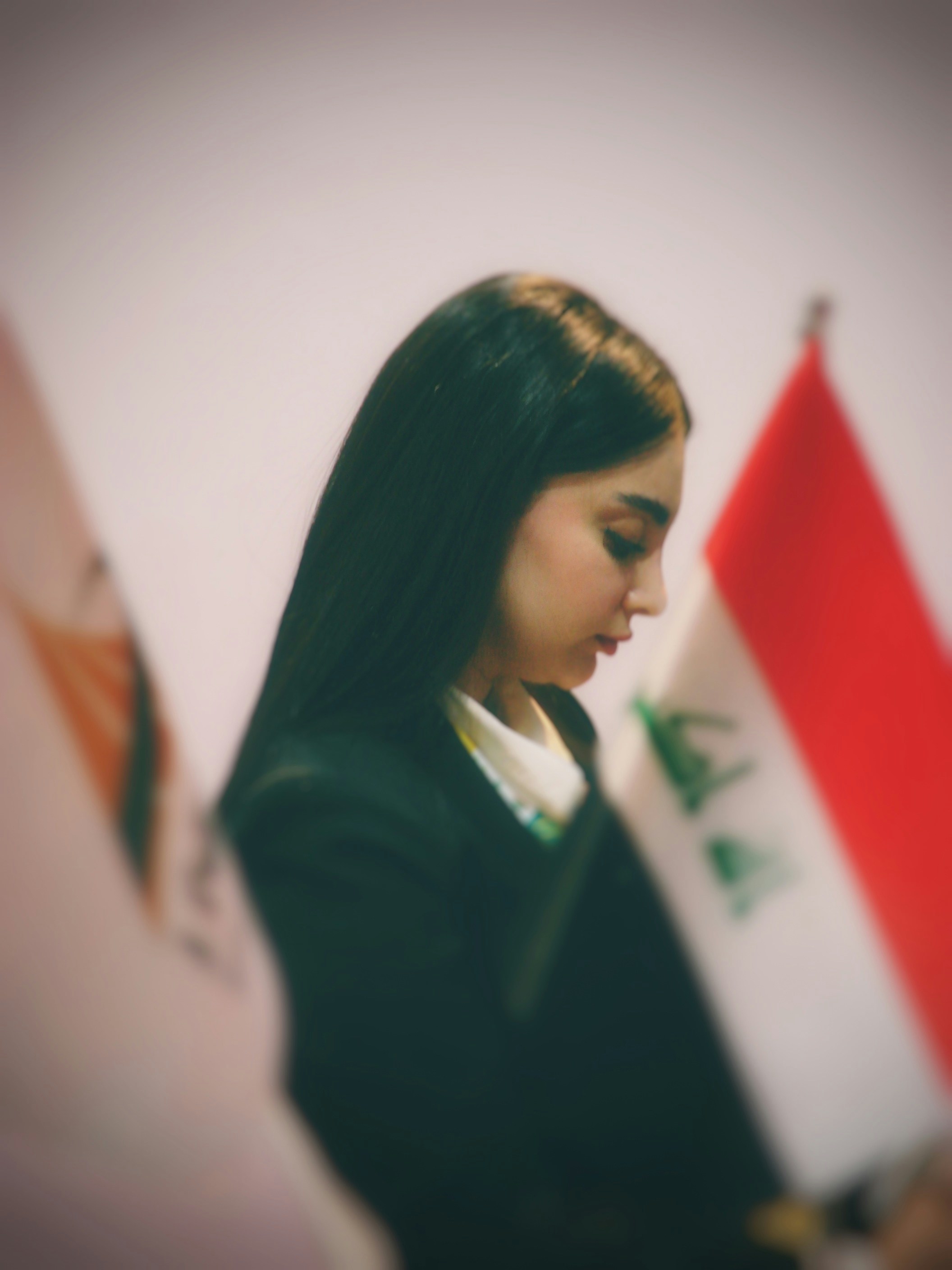 Female figure in formal attire stands beside flags, captured in a shallow depth with soft bokeh showcasing a ceremonial setting.