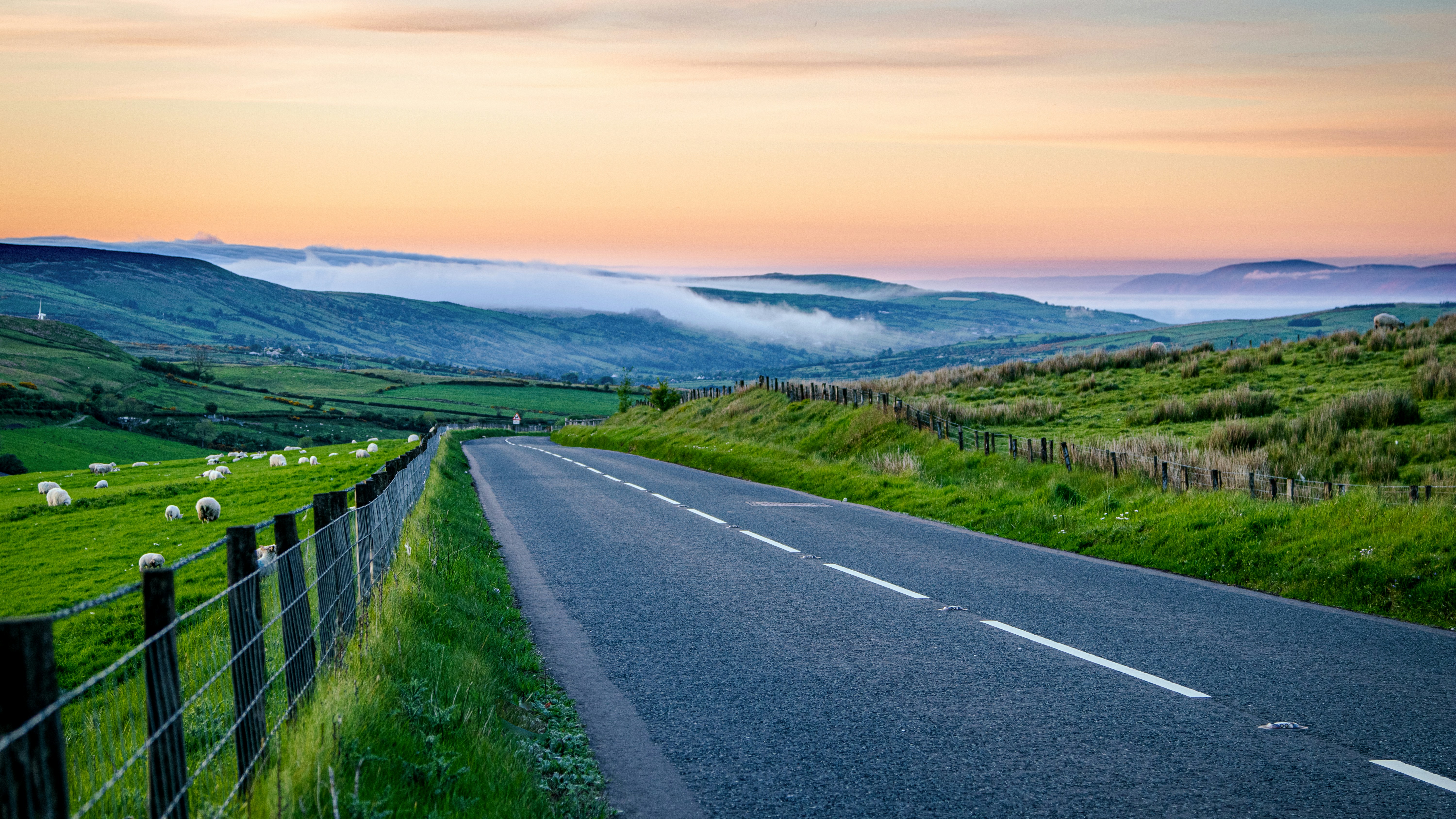 Open rural road winds through green fields with sheep and rolling hills fading into a misty horizon.