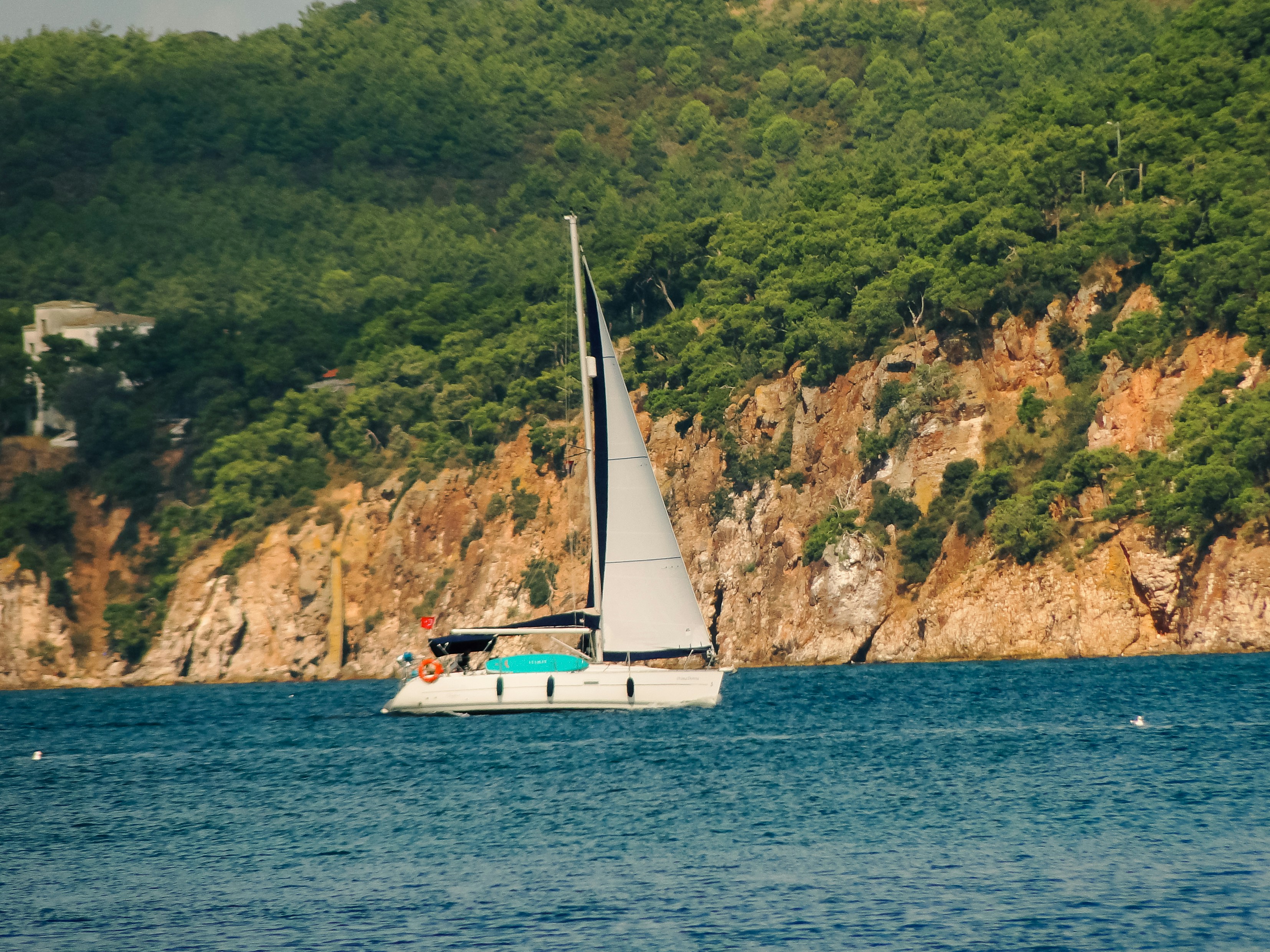 A sailboat with a white sail on blue water near a rocky cliff