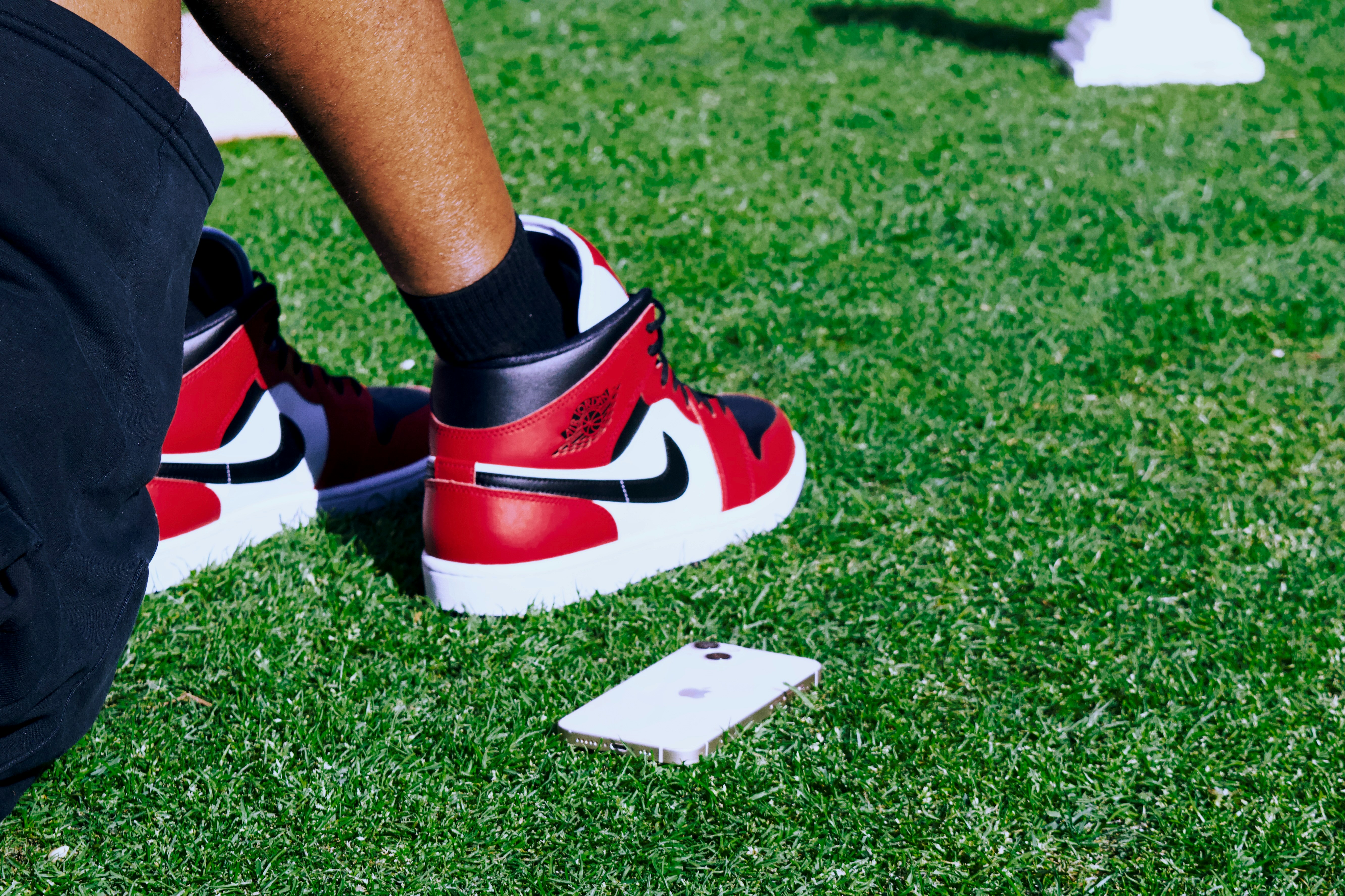 Close-up of a person kneeling on artificial turf with red-and-white sneakers visible, and a white smartphone lying face down nearby; the scene suggests a moment during a practice or drill on a sports field.