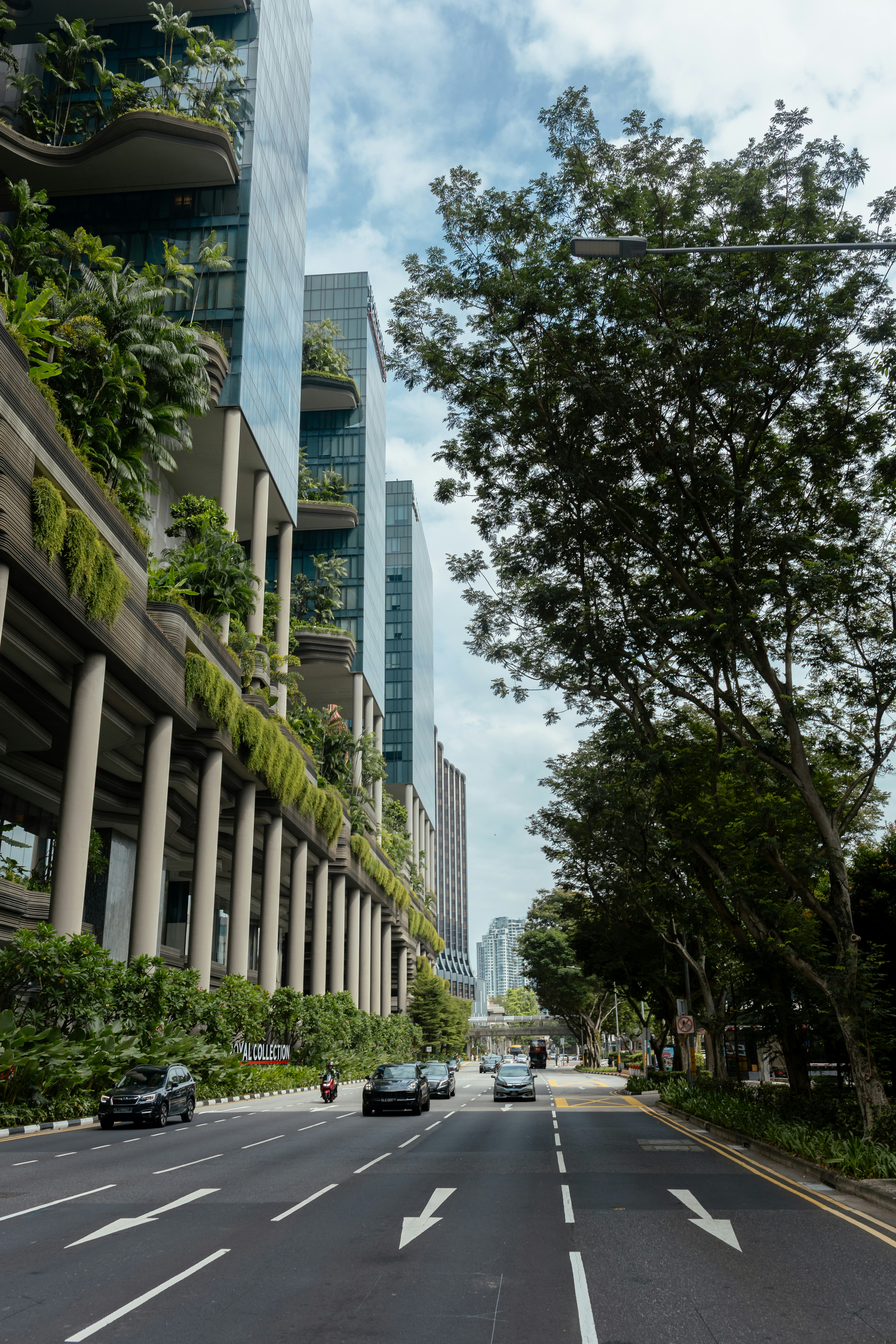 Sunlit city street flanked by a modern glass-and-plant-covered building and tall trees; vehicles travel in multiple lanes beneath a blue sky with scattered clouds.