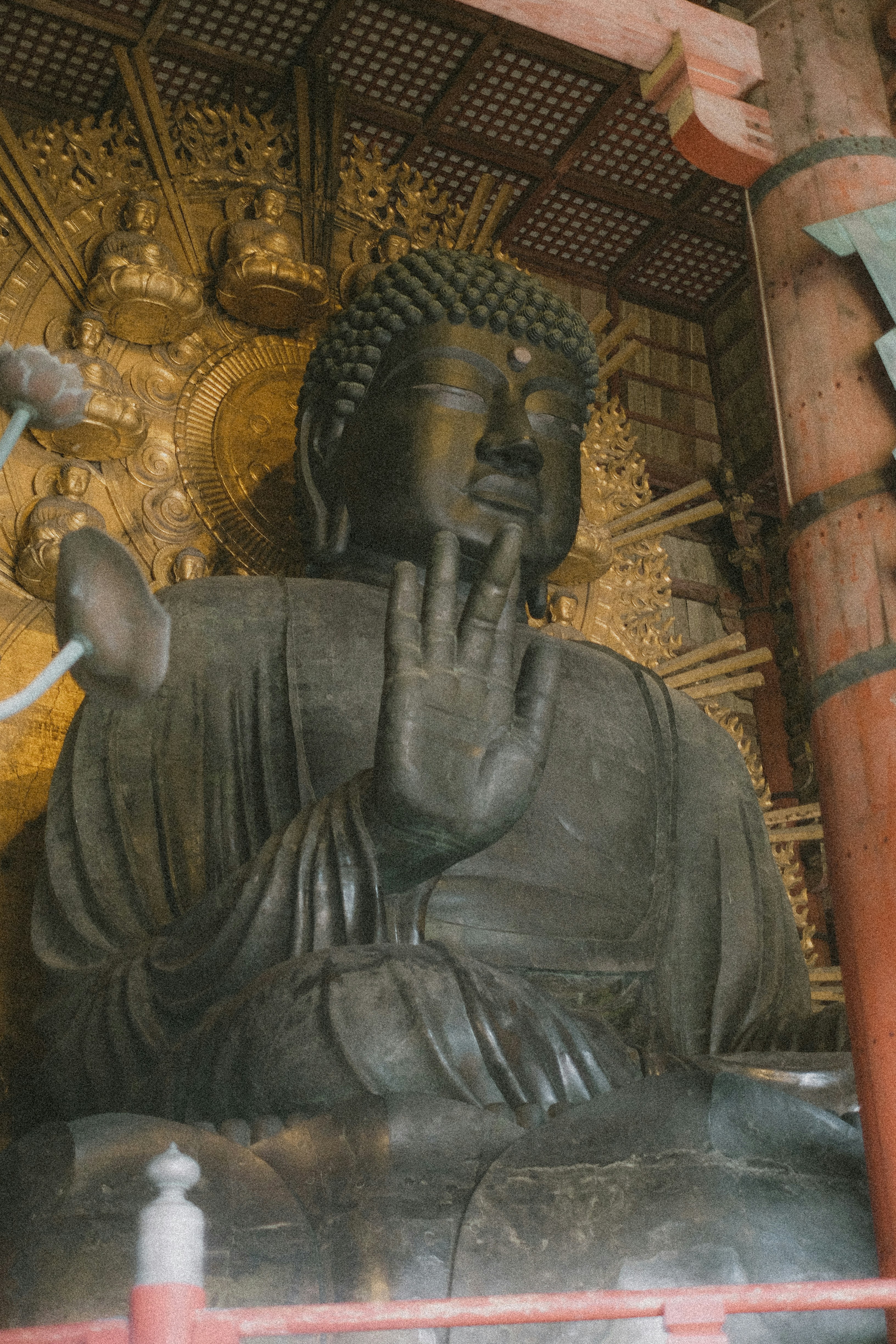 Bronze Buddha statue seated in meditation with a raised palm, set against a gilded backdrop and architectural pillars. The scene conveys tranquil reverence and sacred stillness.