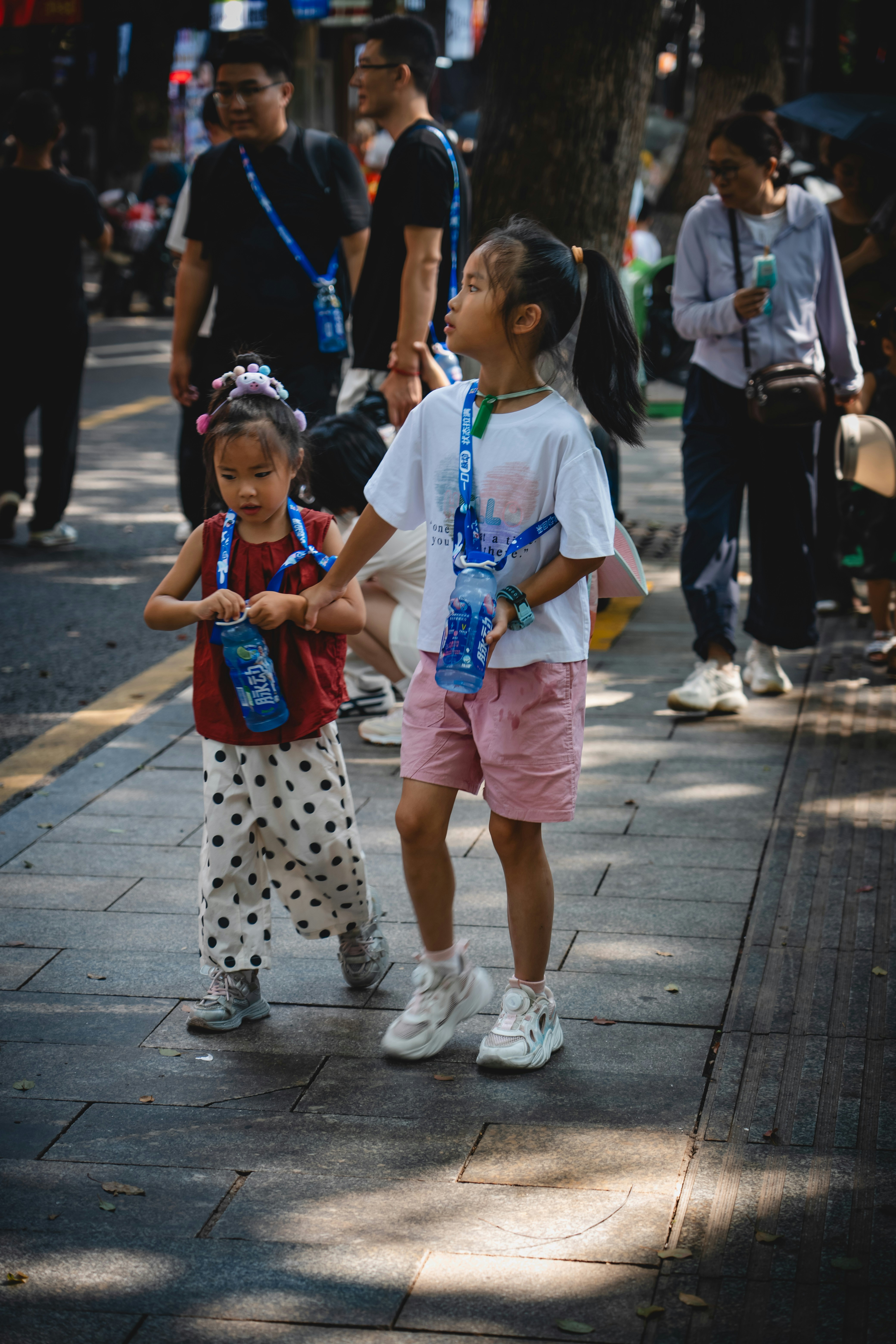 Two Girls in a Crowded Street: Handing Off Bottled Water