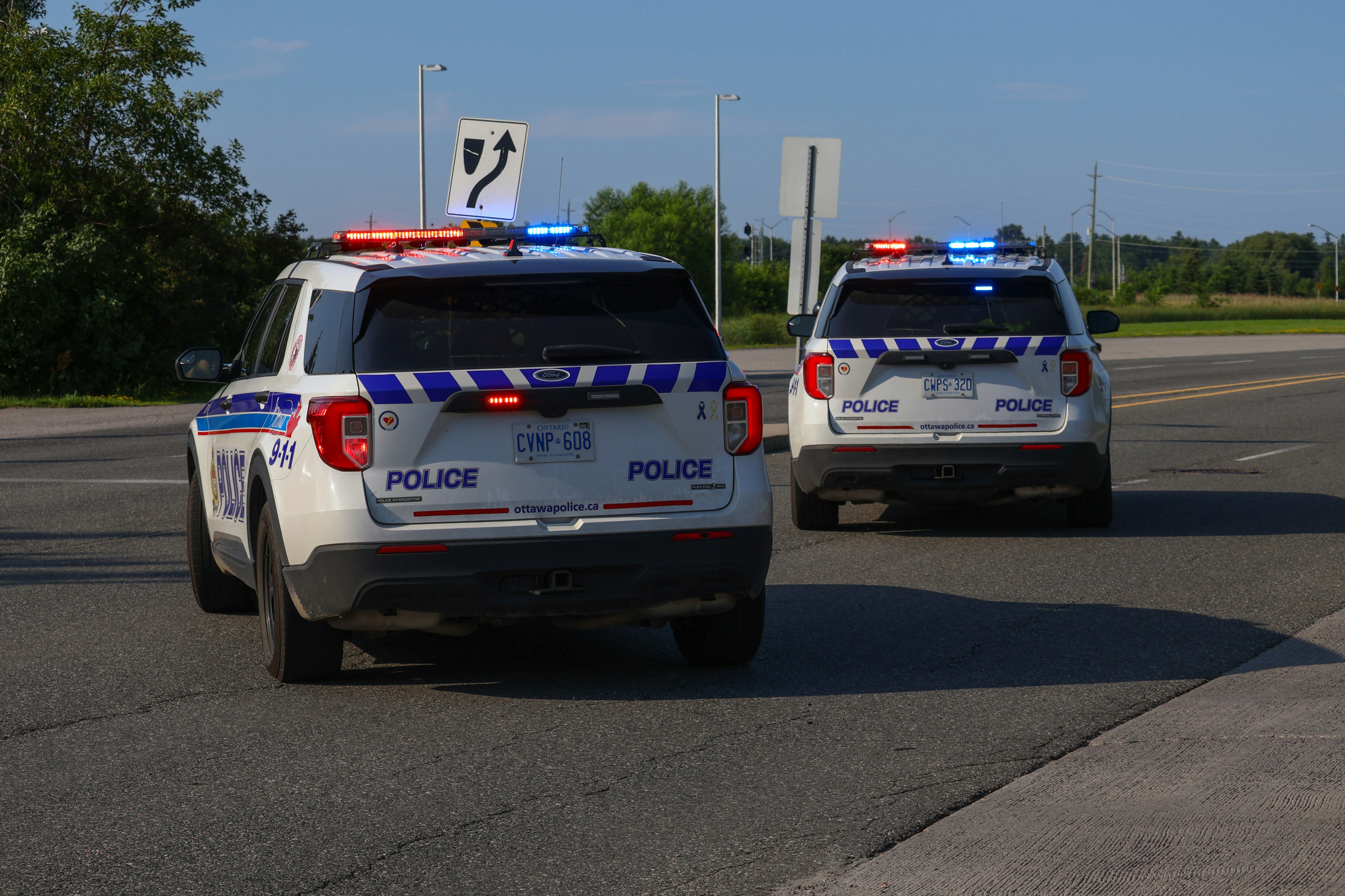 Two police SUVs with illuminated red and blue lights at a rural intersection, seen from behind and in motion on a curved road.