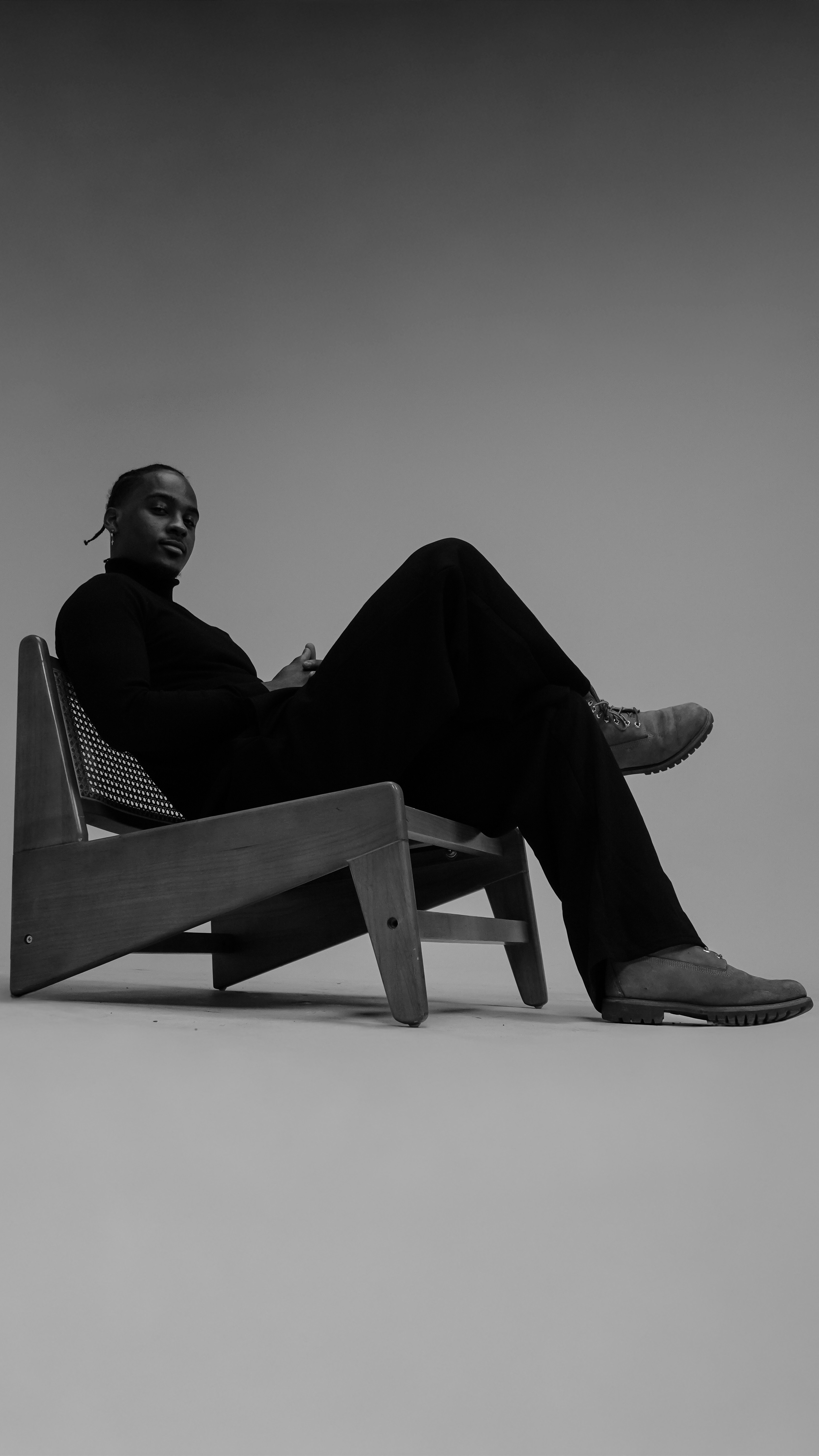 Monochrome portrait of a person lounging in a chair, legs extended, against a seamless backdrop. The image emphasizes relaxed posture and bold silhouette for a contemporary, studio-styled scene.