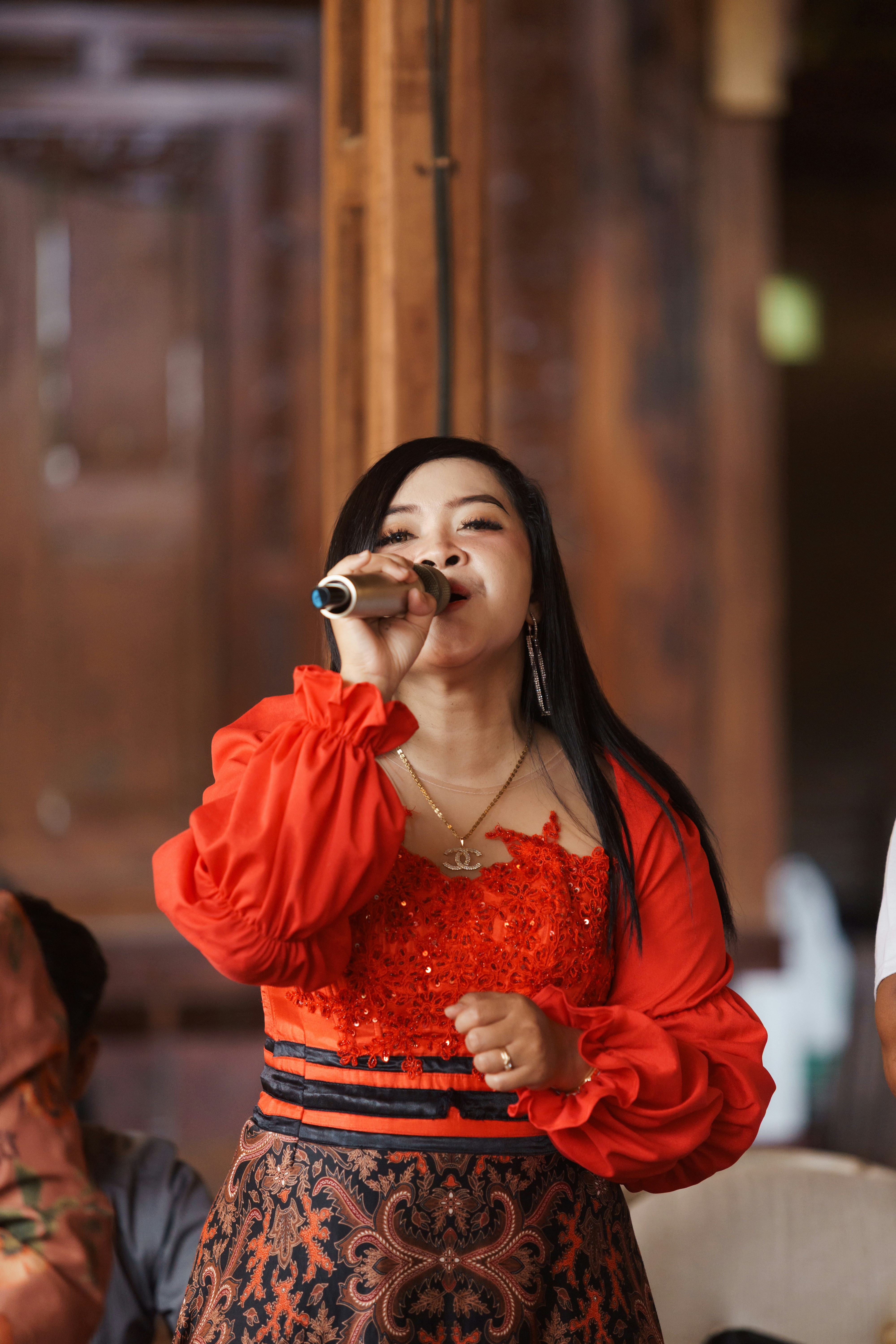 Bold red-sleeved performer singing into a cordless mic, stage setting with wooden backdrop and warm tones.