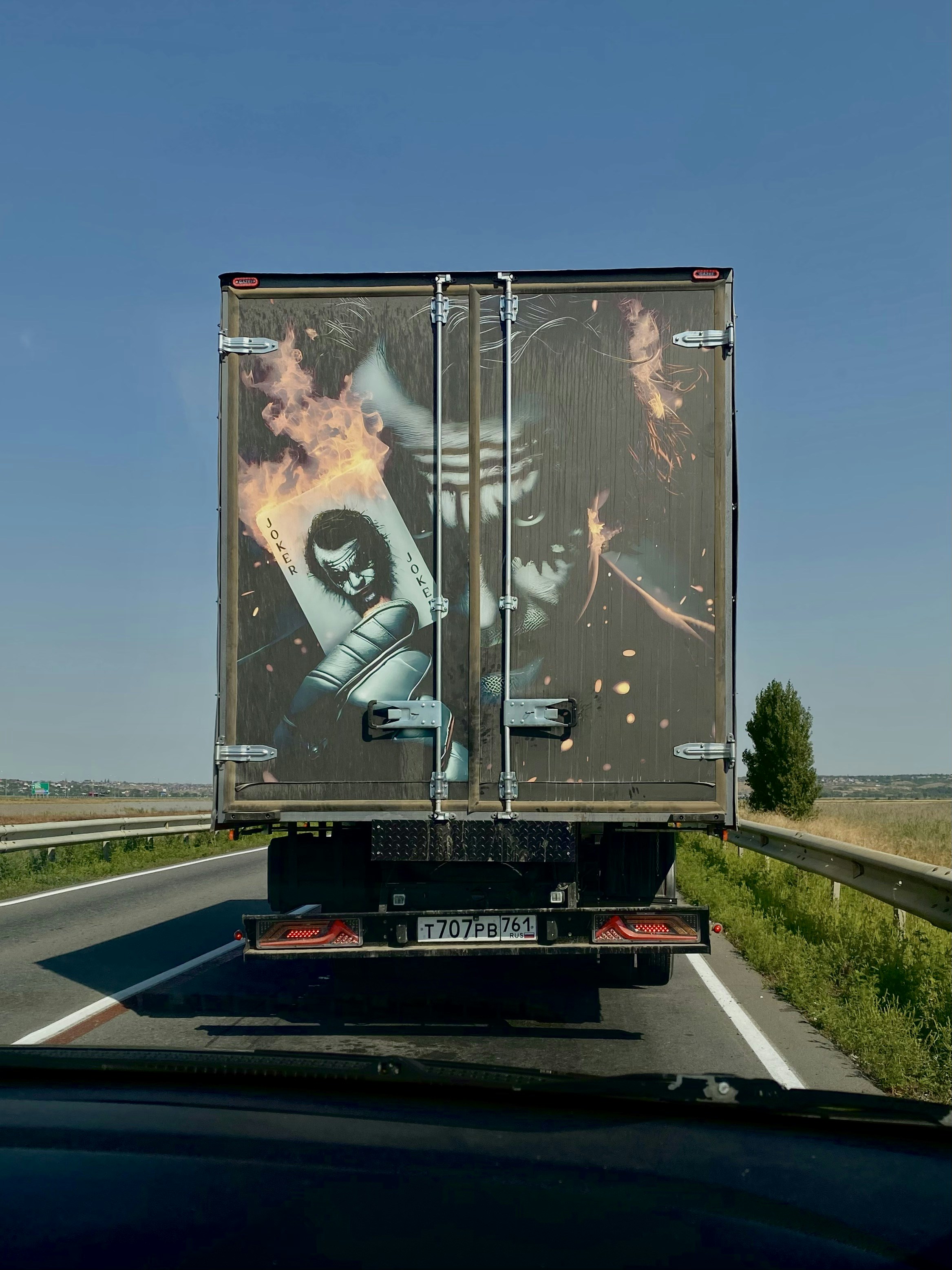 A trailer with a graphic face on its back doors is viewed from inside a car on a straight highway, illuminated by daylight; the scene captures motion and roadside scenery with clear sky and distant fields.