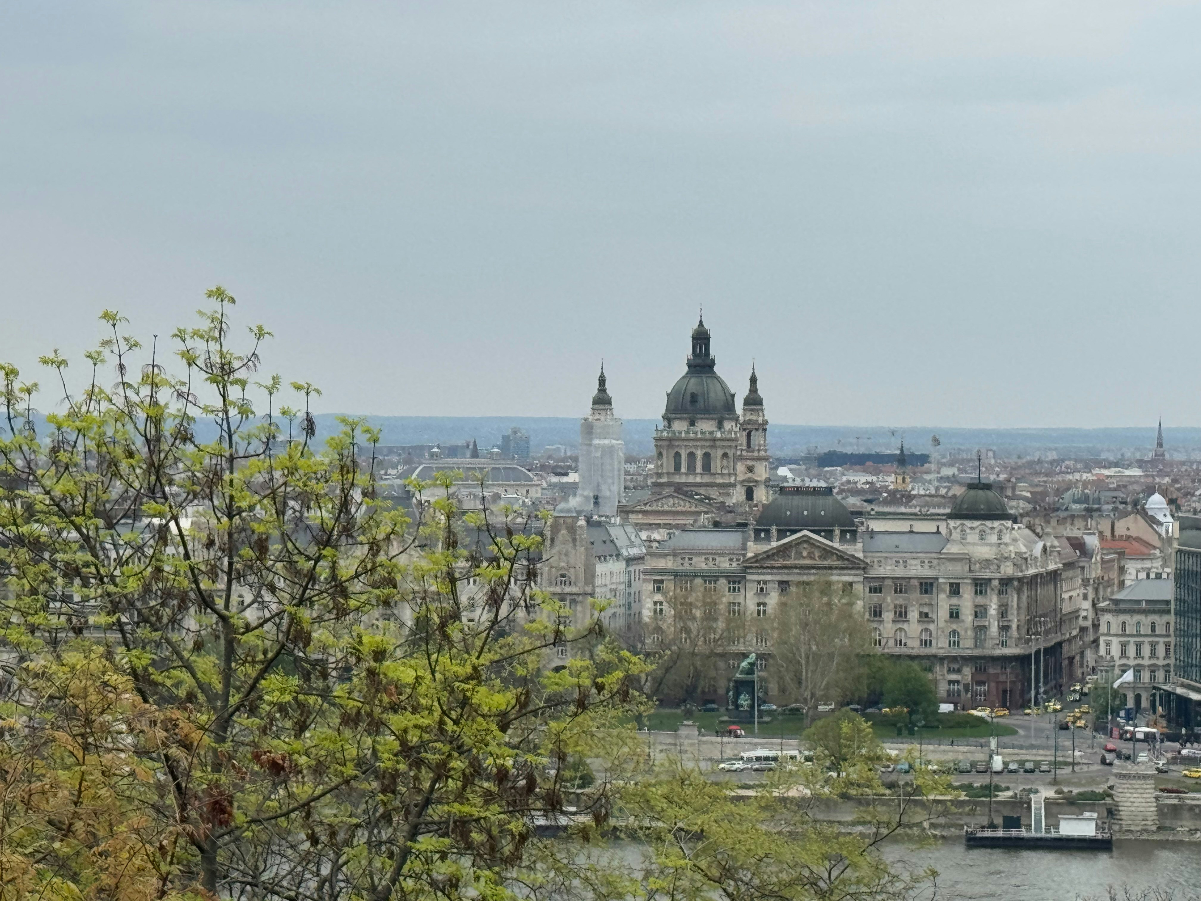 Urban panorama featuring a historic domed building rising above a mid-distance street scene, framed by foreground trees; a calm, overcast sky sets a muted mood. (Logo/illustration: none)