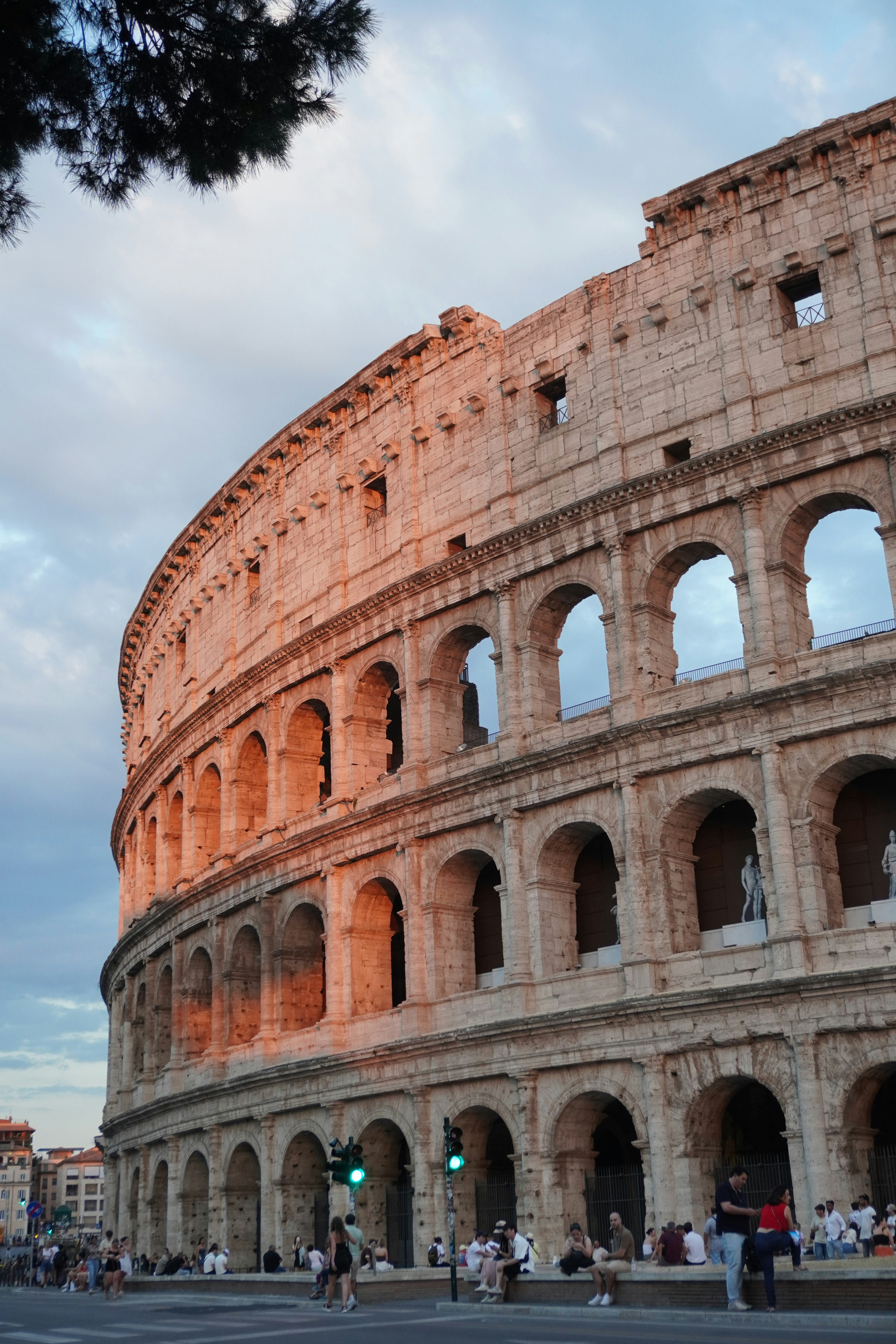 Ancient Roman Colosseum facade bathed in warm sunset light with people gathered along the base under a pale blue sky.