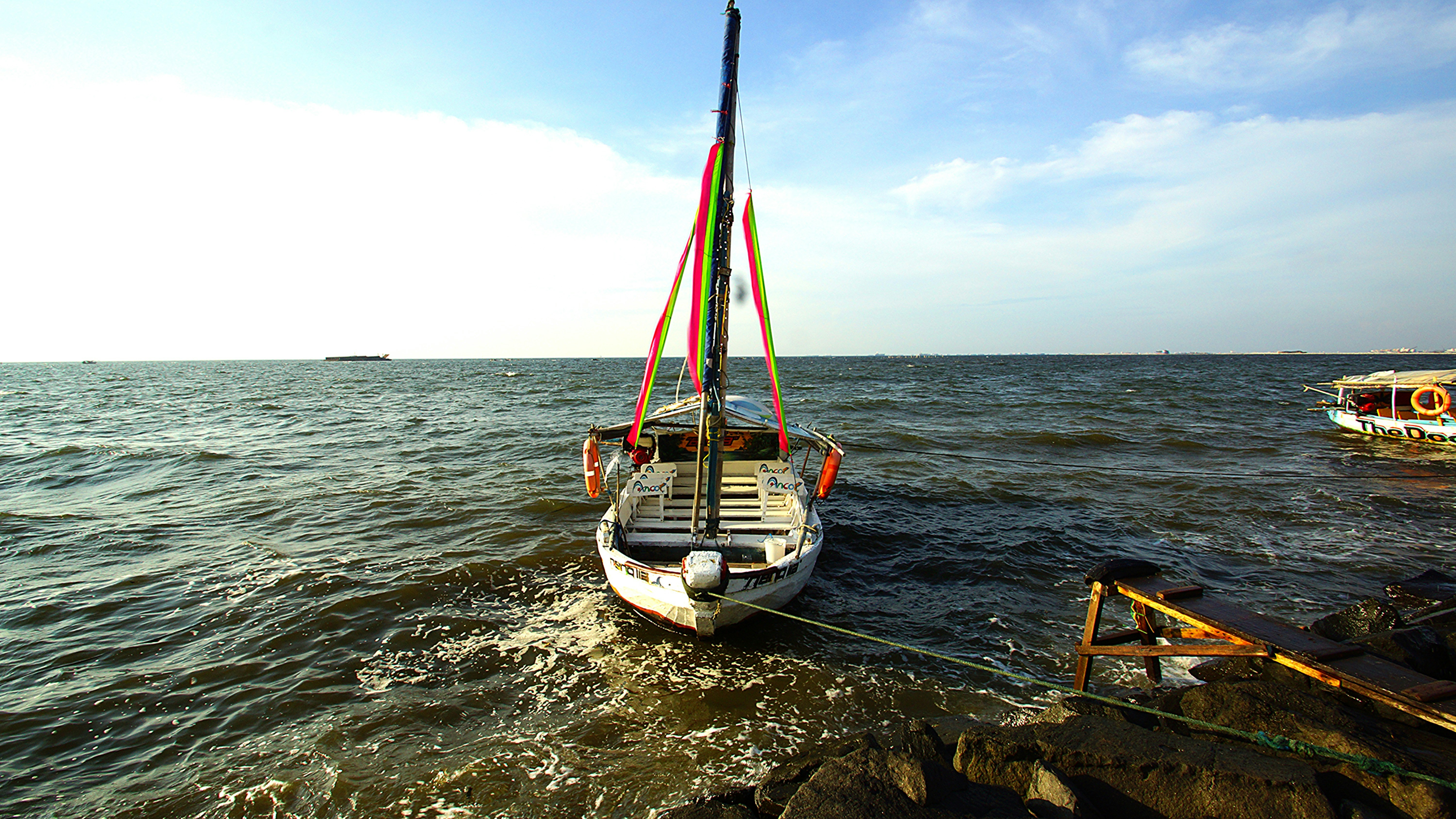 A small sailboat anchored near a rocky breakwater with colorful pennants fluttering in a calm sea under a clear sky.