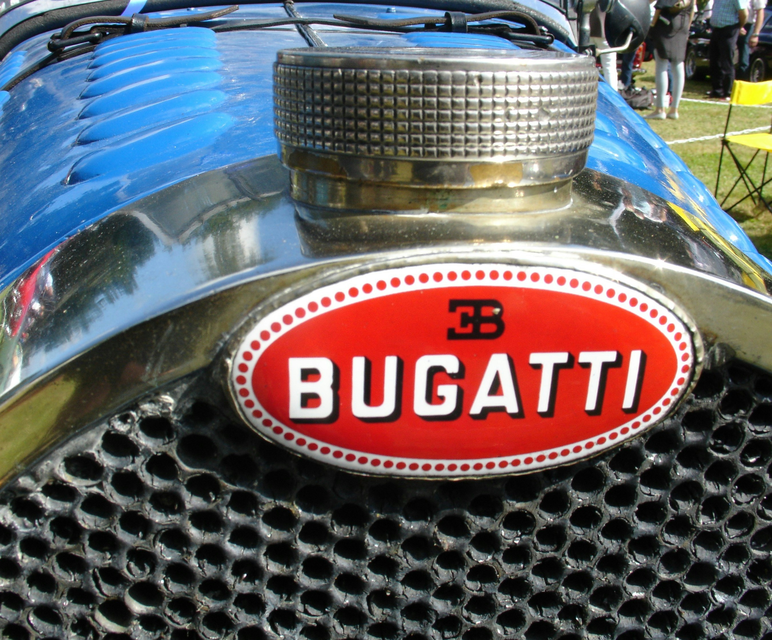 Chrome Bugatti badge centered above a honeycomb radiator grille on a vintage blue car. Clear, glossy surfaces reflect the surrounding scene, suggesting an outdoor car show setting.