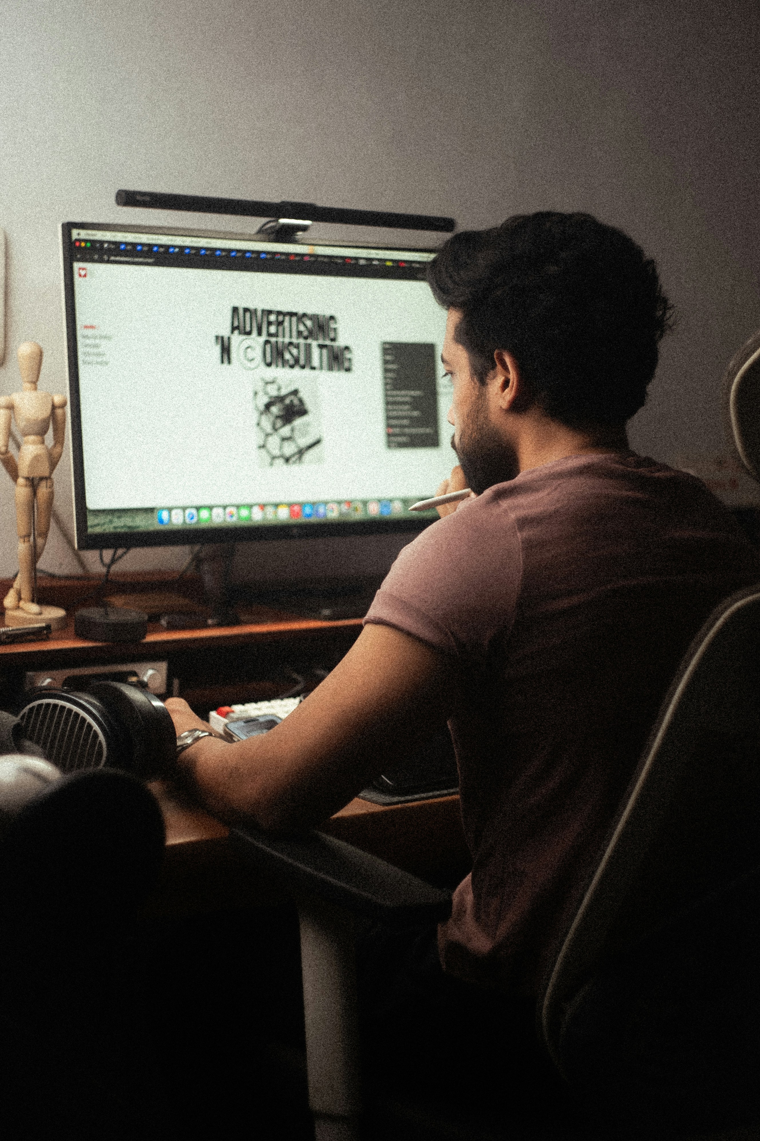 A man sits at a desk facing a computer monitor displaying a design page, with studio tools and a wooden mannequin on the left; a headphone sits in front of the viewer. This is a workspace photo.