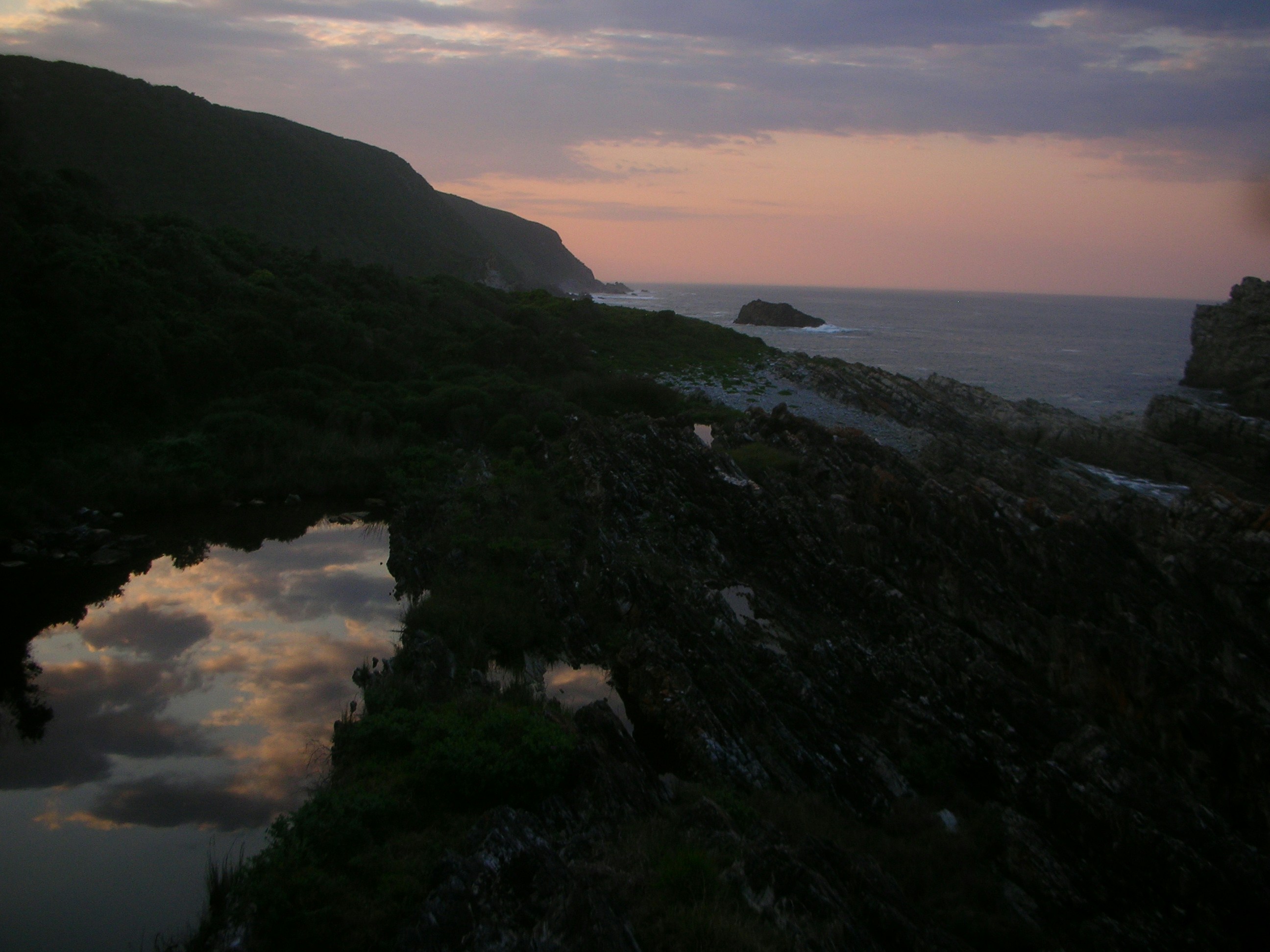 Dusk lights the rocky coastline with calm pools reflecting the sky; rugged foreground leads to a distant horizon by the sea. This landscape emphasizes mood and texture, captured as a natural scene.
