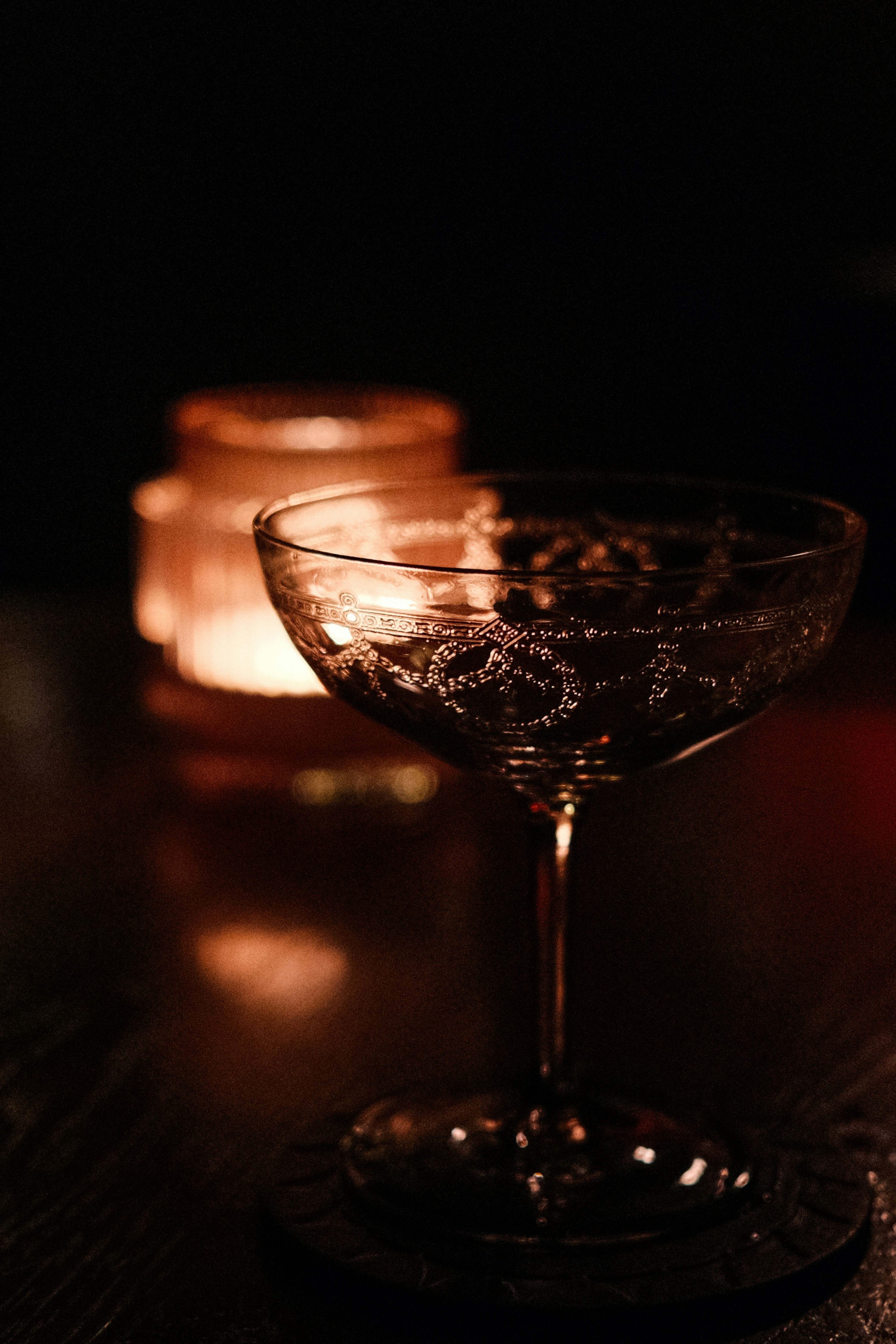 A coupe glass with an ornate lace pattern sits on a dark surface, illuminated by a warm candle glow in the background.