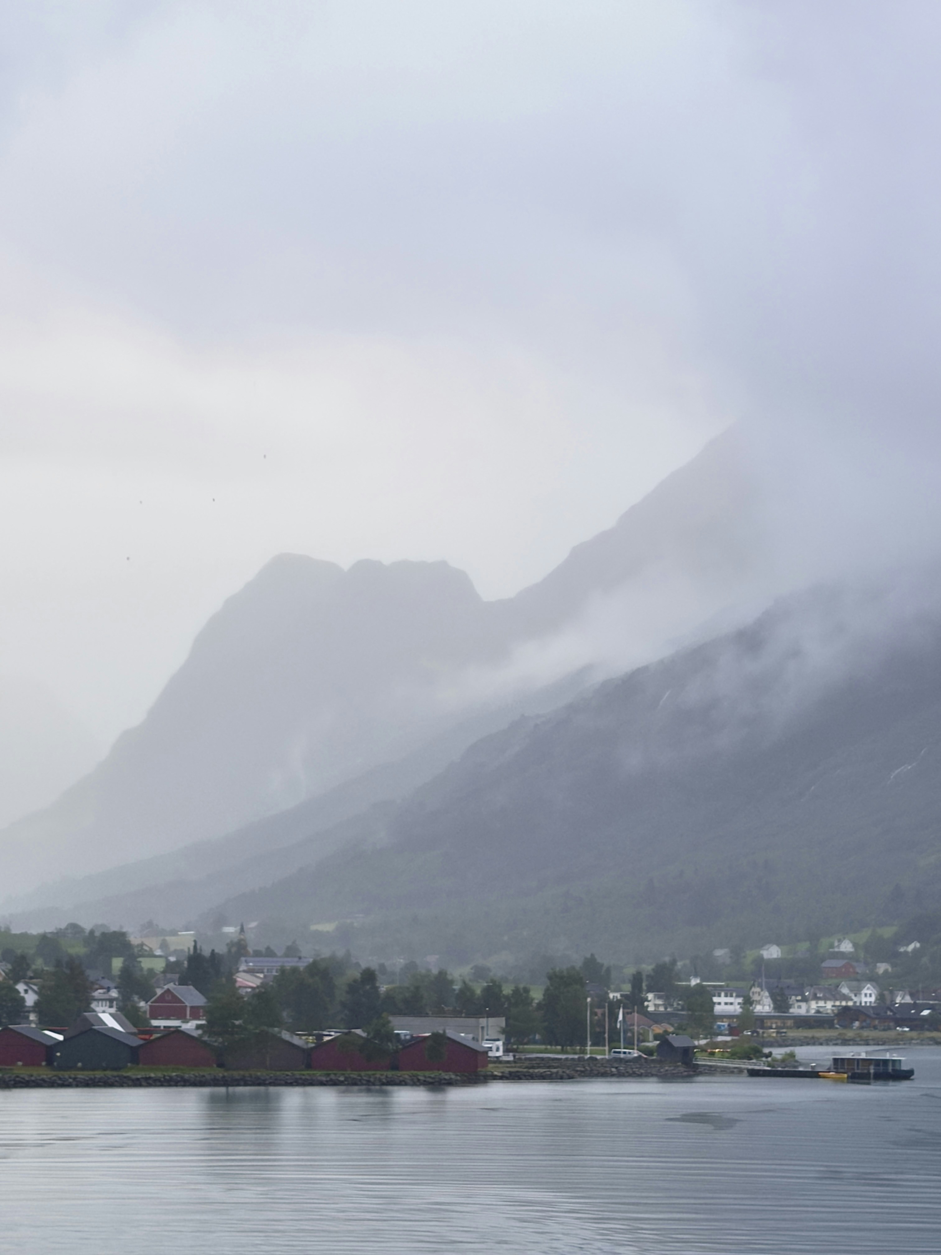 Low-lying village along calm water with misty mountains rising in the background, enveloped in soft fog.
