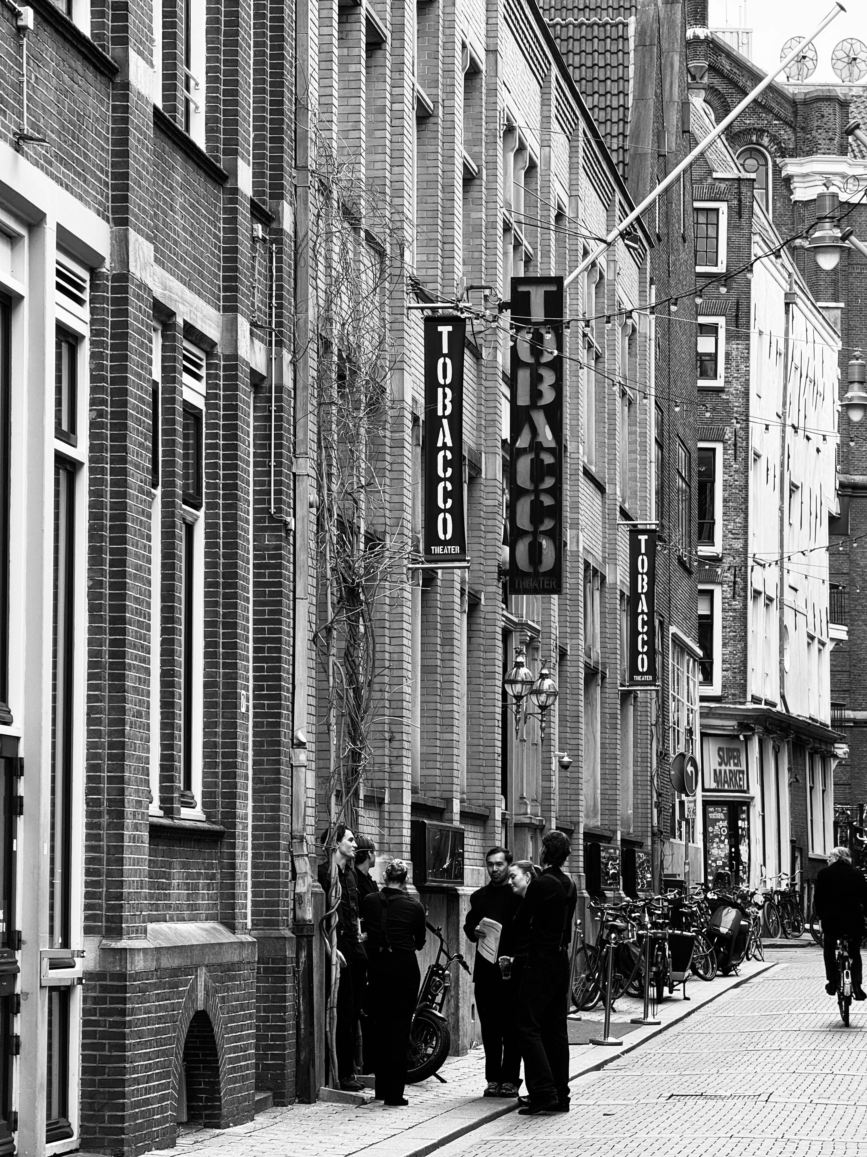 A monochrome city street scene with brick buildings, hanging tobacco shop signs, and a small cluster of people near the curb, captured in candid street photography style.