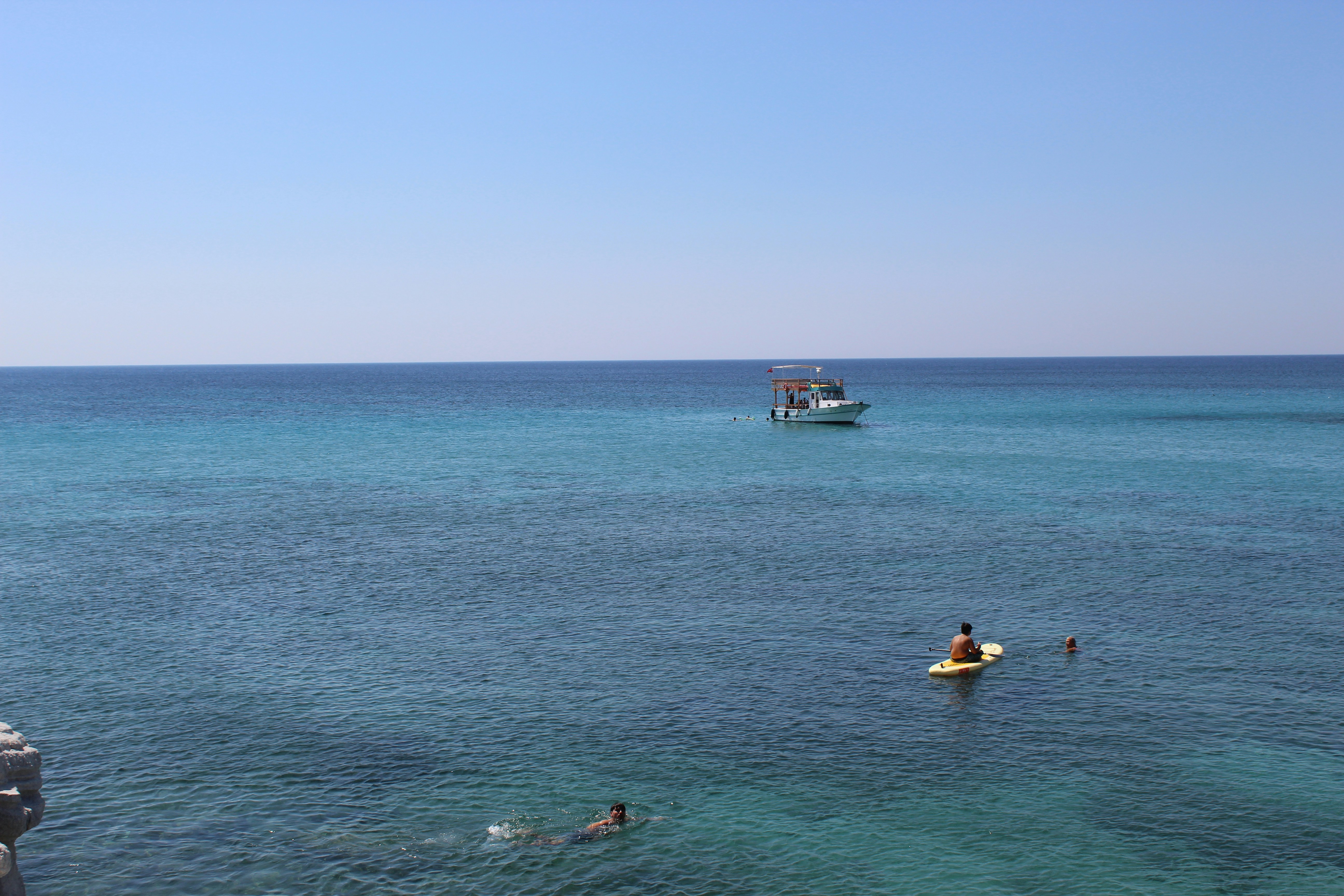 Calm tropical sea with a small boat near the horizon and a bright swimmer in a yellow kayak close to the right foreground; the scene emphasizes tranquil water and distant horizon in a sunny coastal setting.