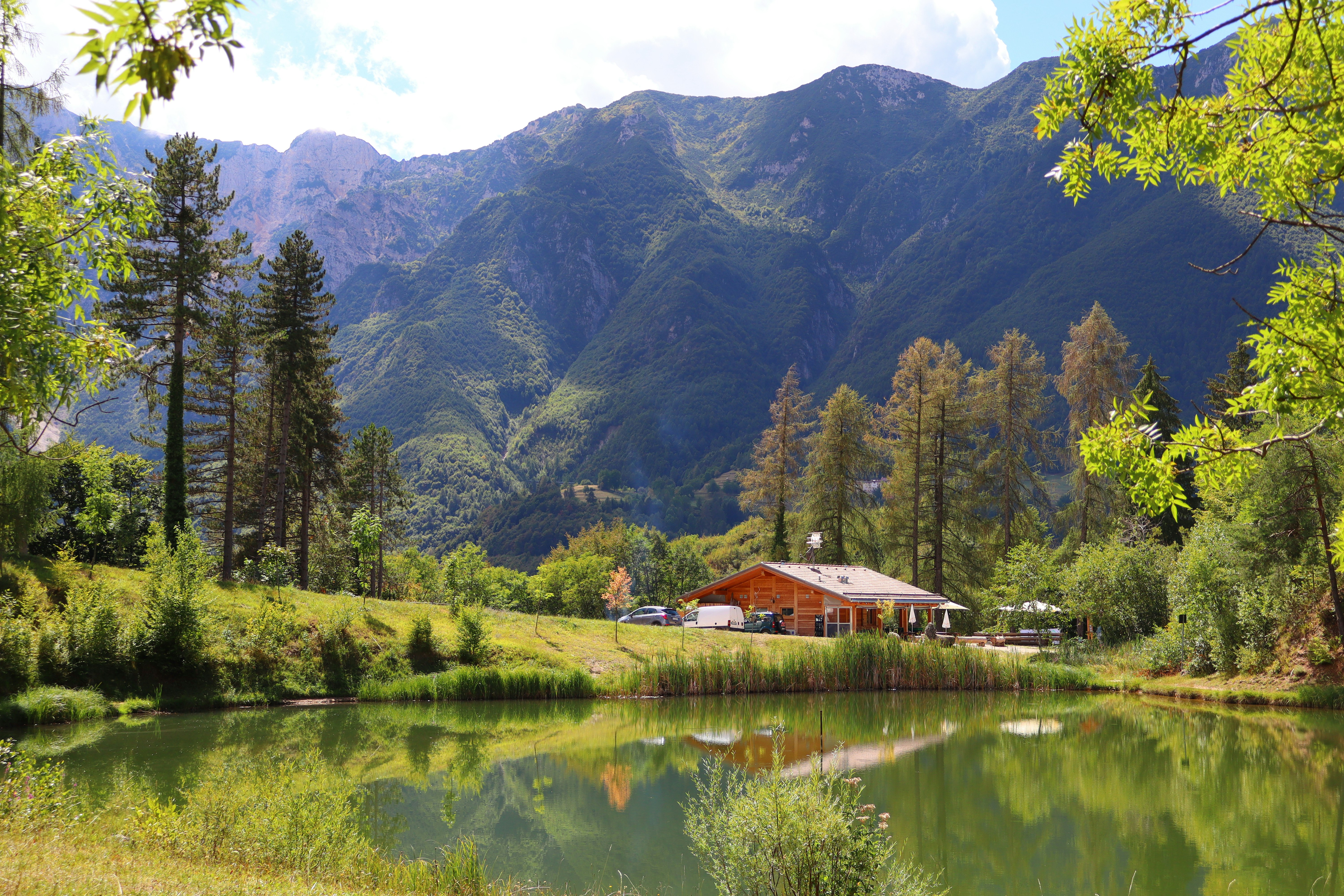 A tranquil alpine lodge sits beside a reflective green lake with pine trees and towering mountains in the background.