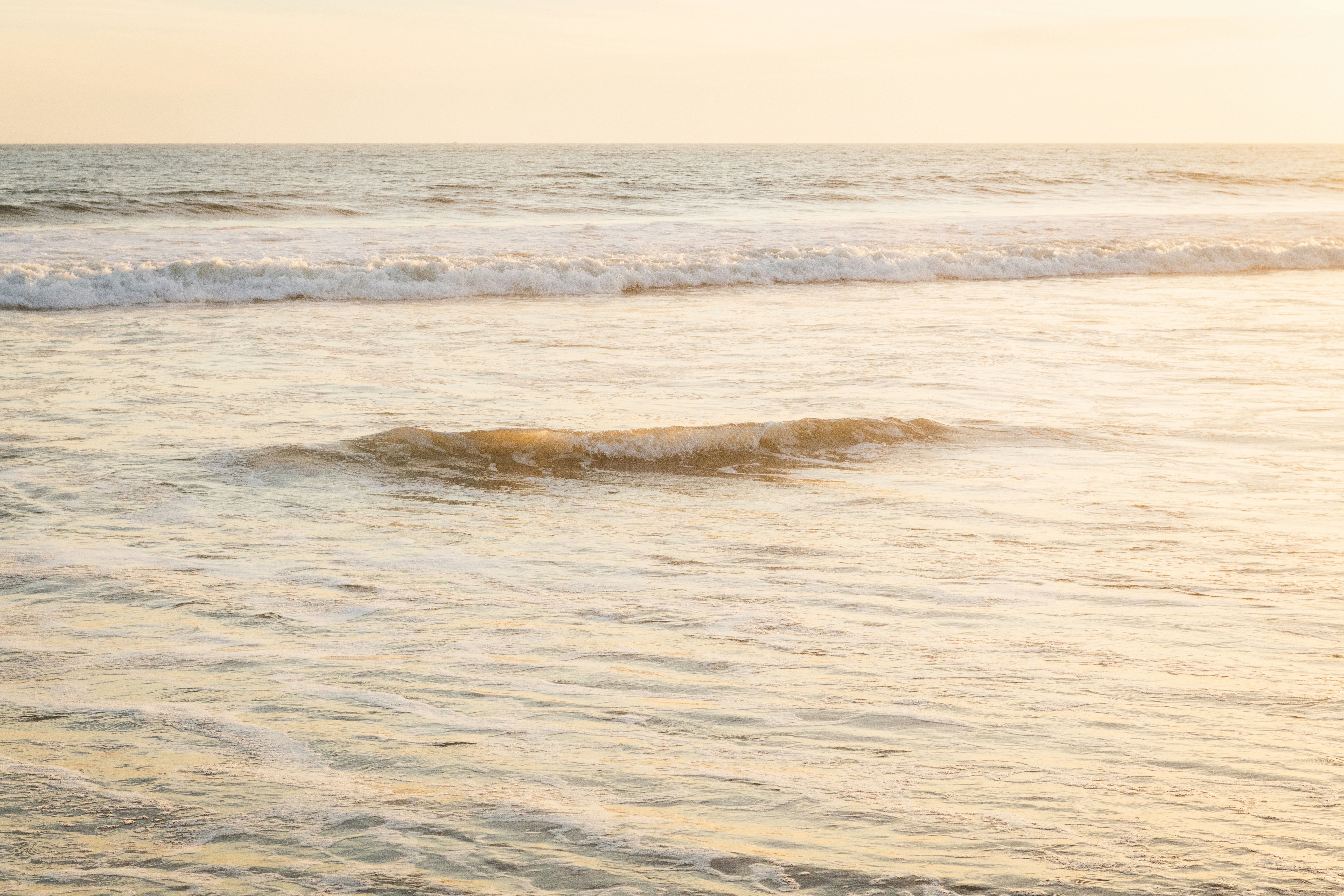 Soft, sunlit shoreline with gentle waves rolling in at sunset, a calm seascape emphasizing warm tones and expansive horizon.