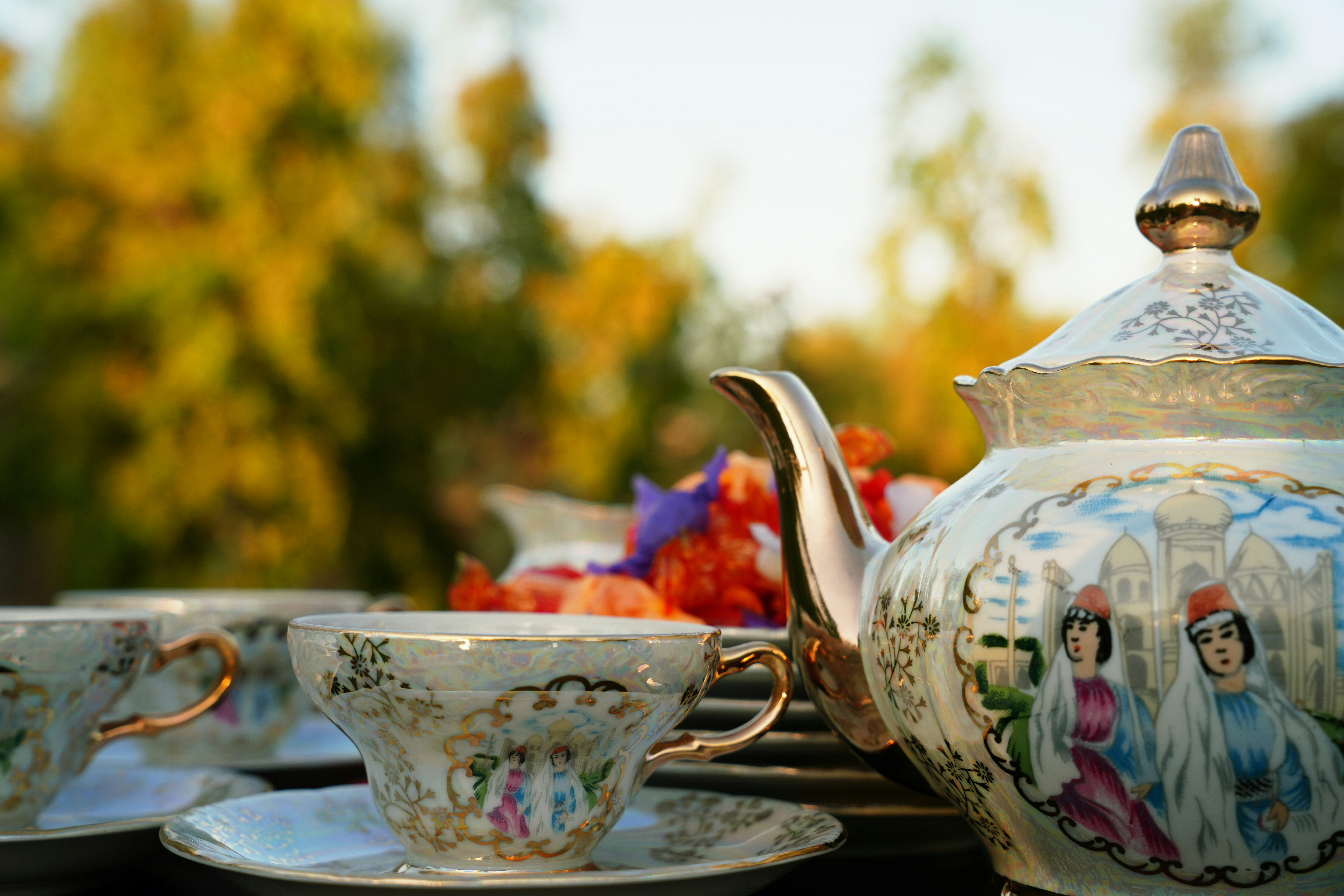 Delicate porcelain teacups and a matching teapot sit on a tray, catching warm afternoon light with a soft, blurred outdoor backdrop.