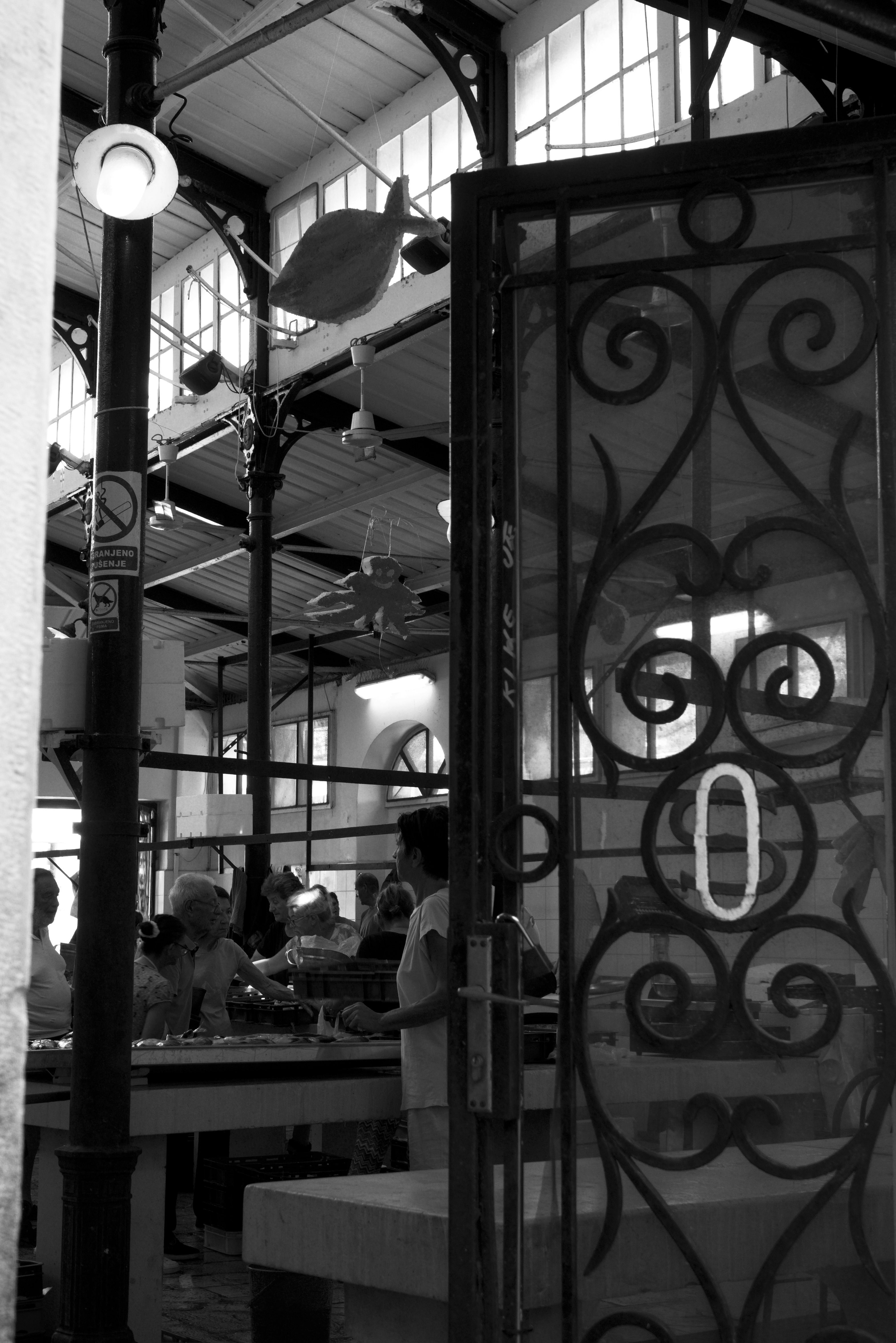 A grayscale scene inside a bustling market hall through an ornate door, with people gathered around a counter and architectural ironwork framing the doorway.