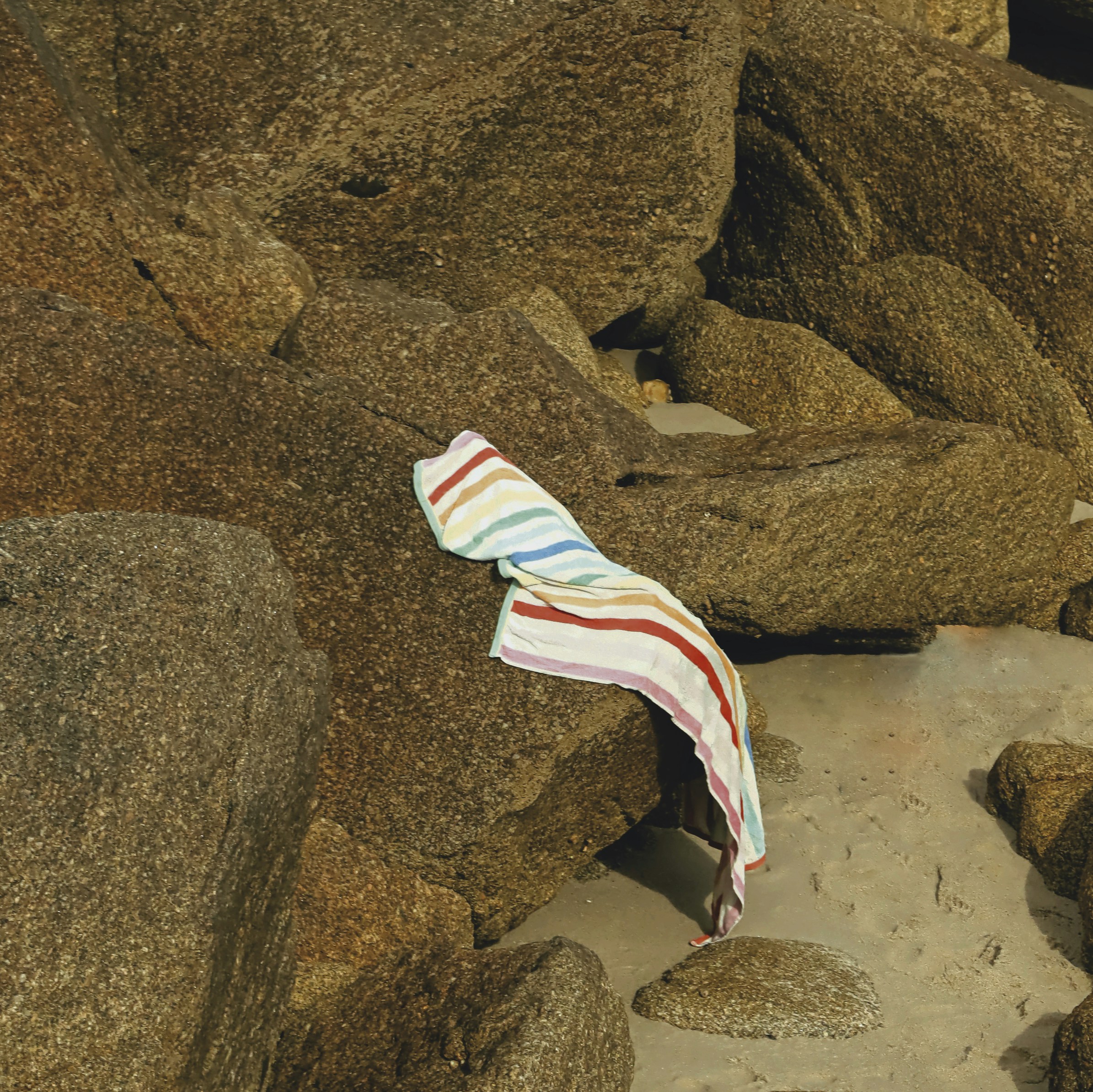 A striped towel lies draped over sunlit rocks with sand at the base, suggesting a tranquil beach scene and a moment of pause.