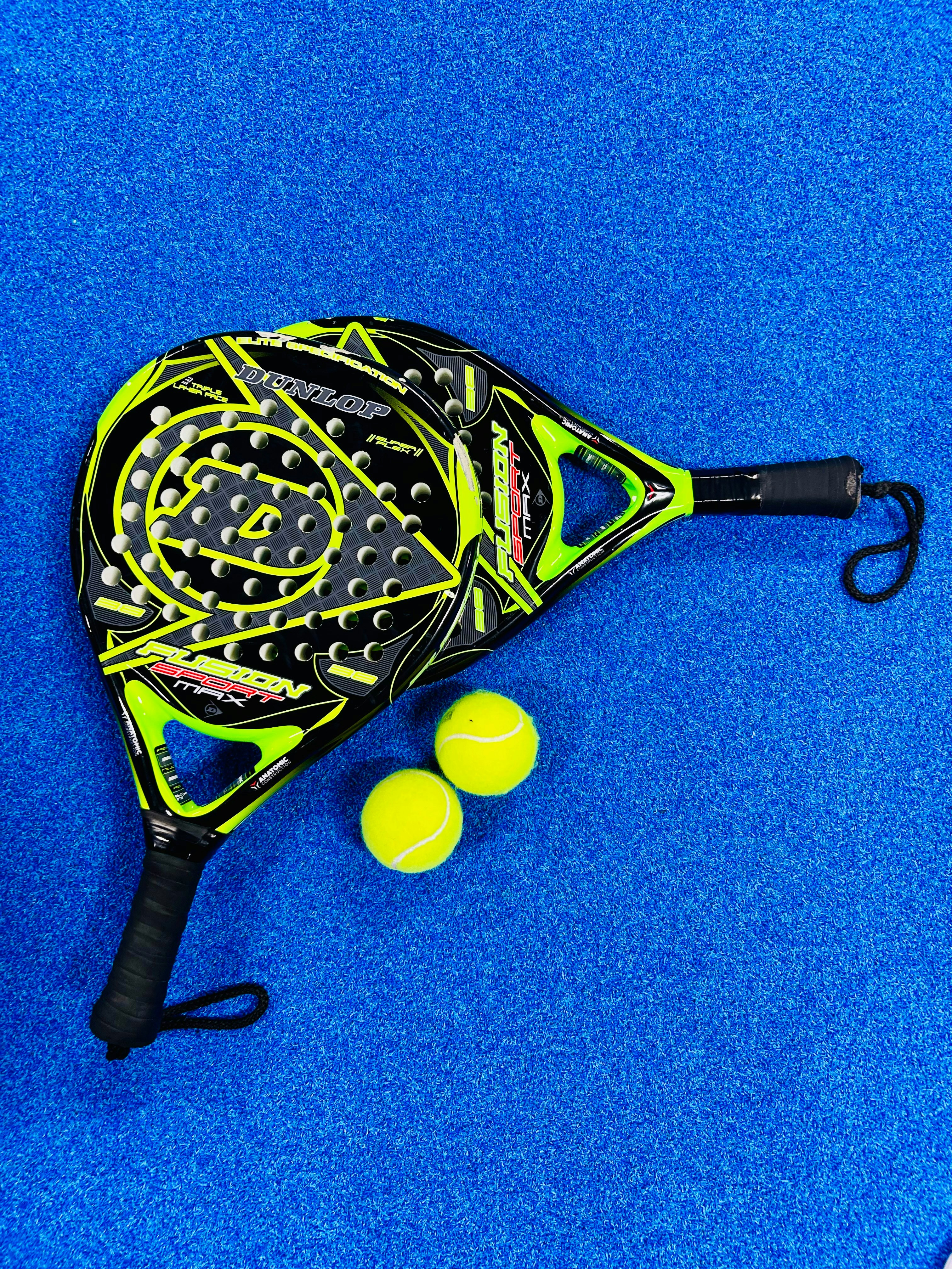 Bright yellow tennis balls lie beside a neon-accent paddle on a blue court surface.