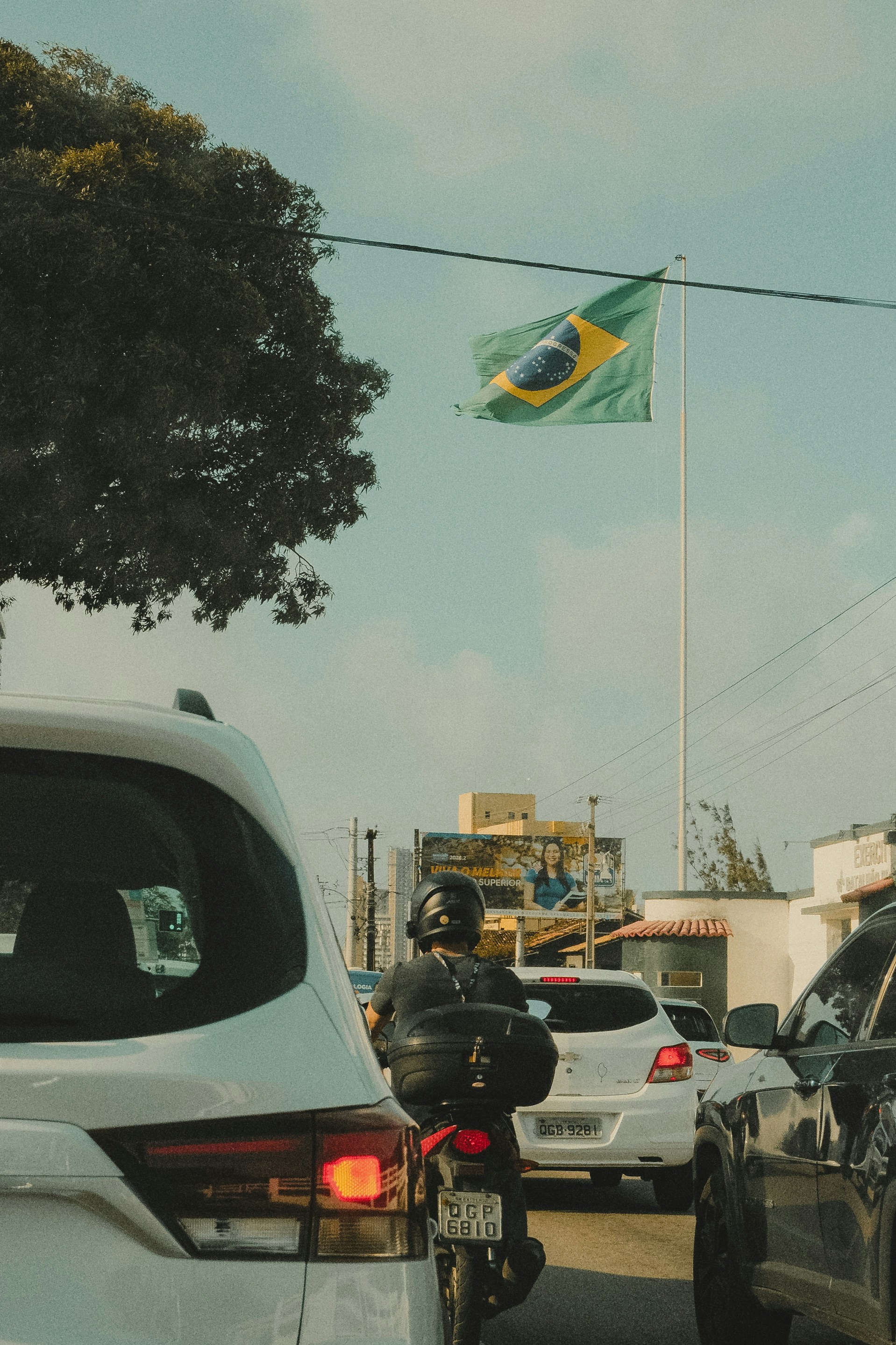 A motorcyclist in traffic beneath a large Brazilian flag on a tall pole