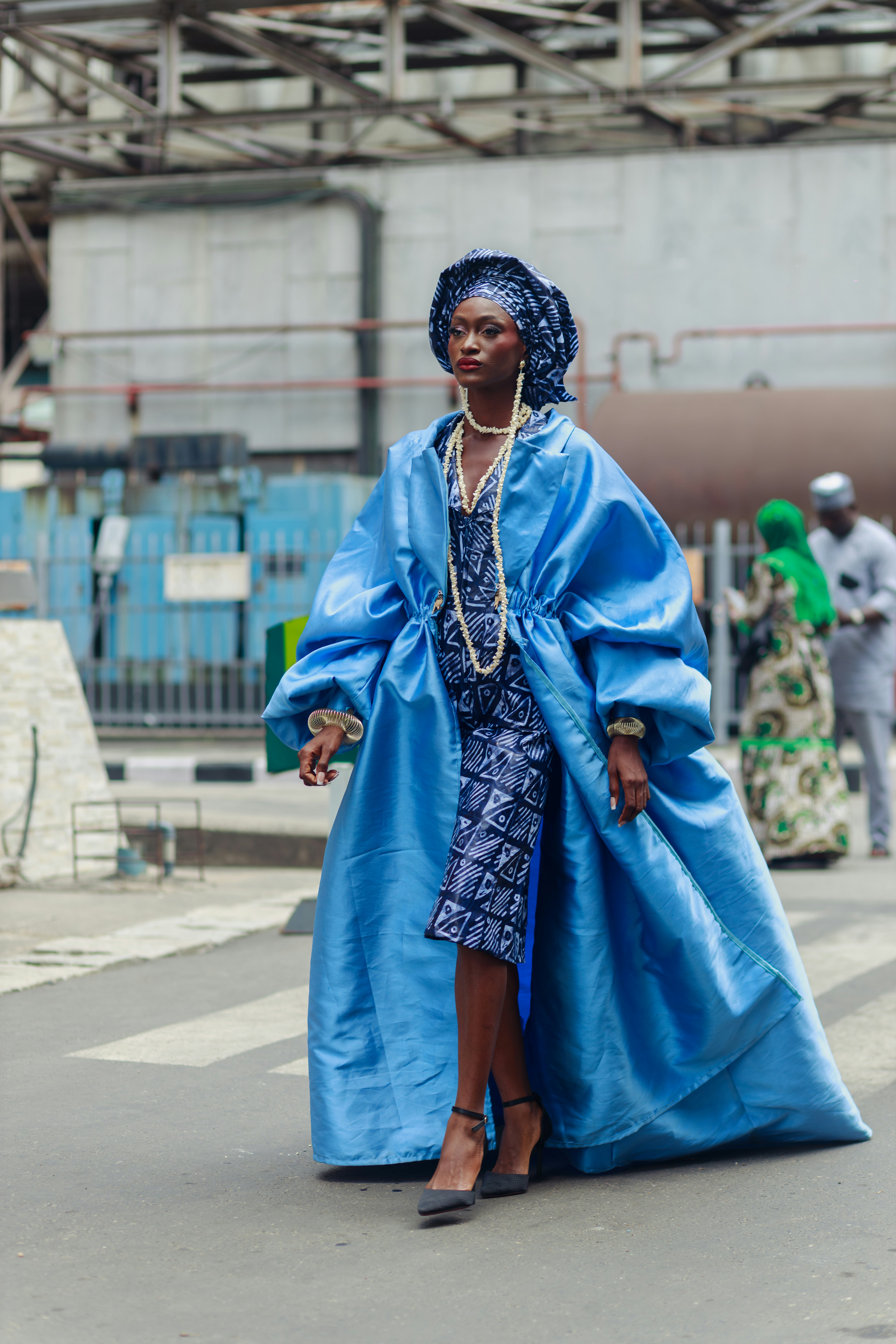 A striking woman in a voluminous blue coat and patterned dress walks confidently down an urban street, accessorized with a beaded headwrap and pearls. The scene captures a fashion-forward moment amid industrial backdrop.