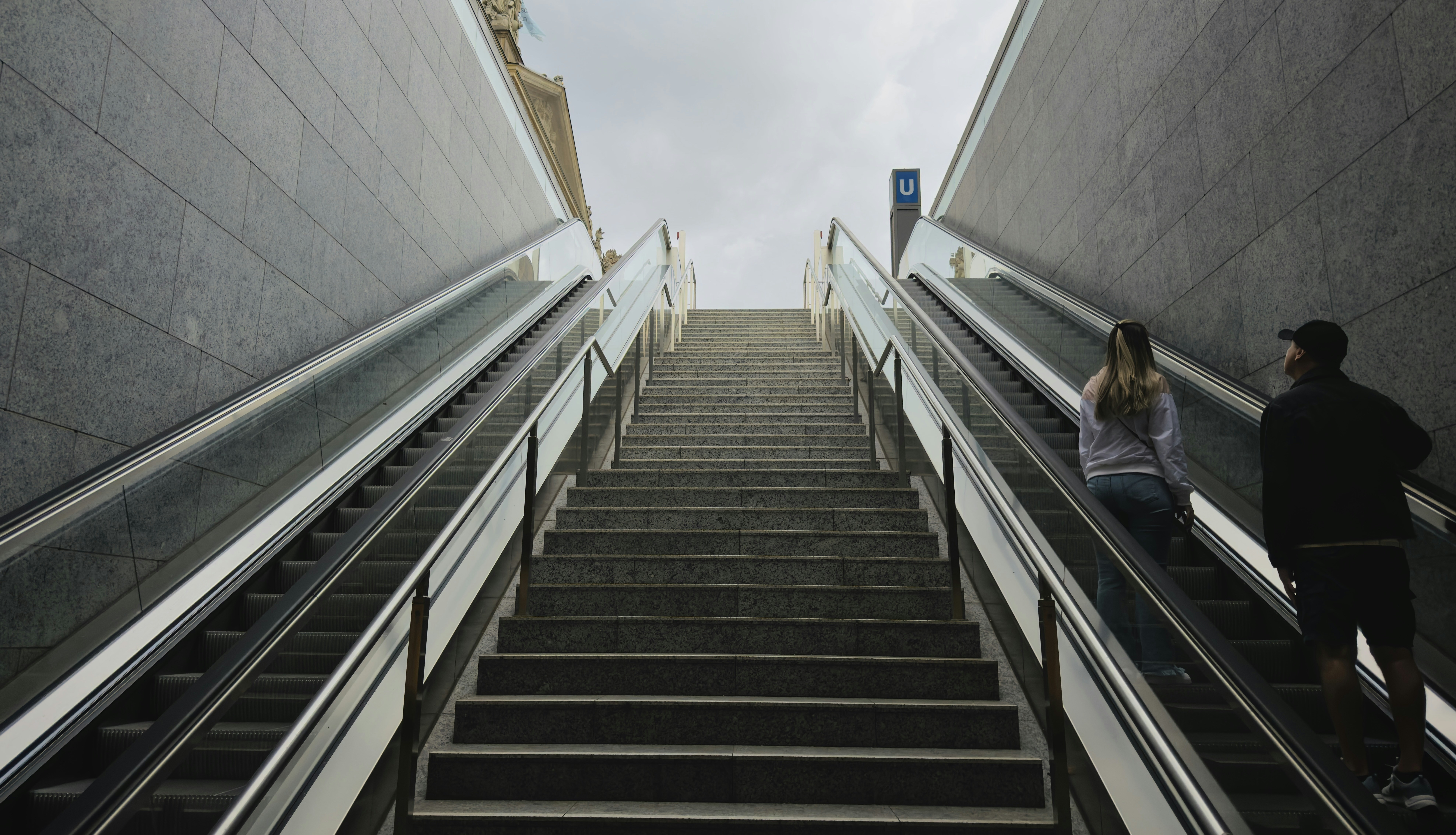 Escalator stairs leading upward between stone walls, two commuters on the right side moving up, soft daylight from above. A clean, architectural moment with a hint of human motion.