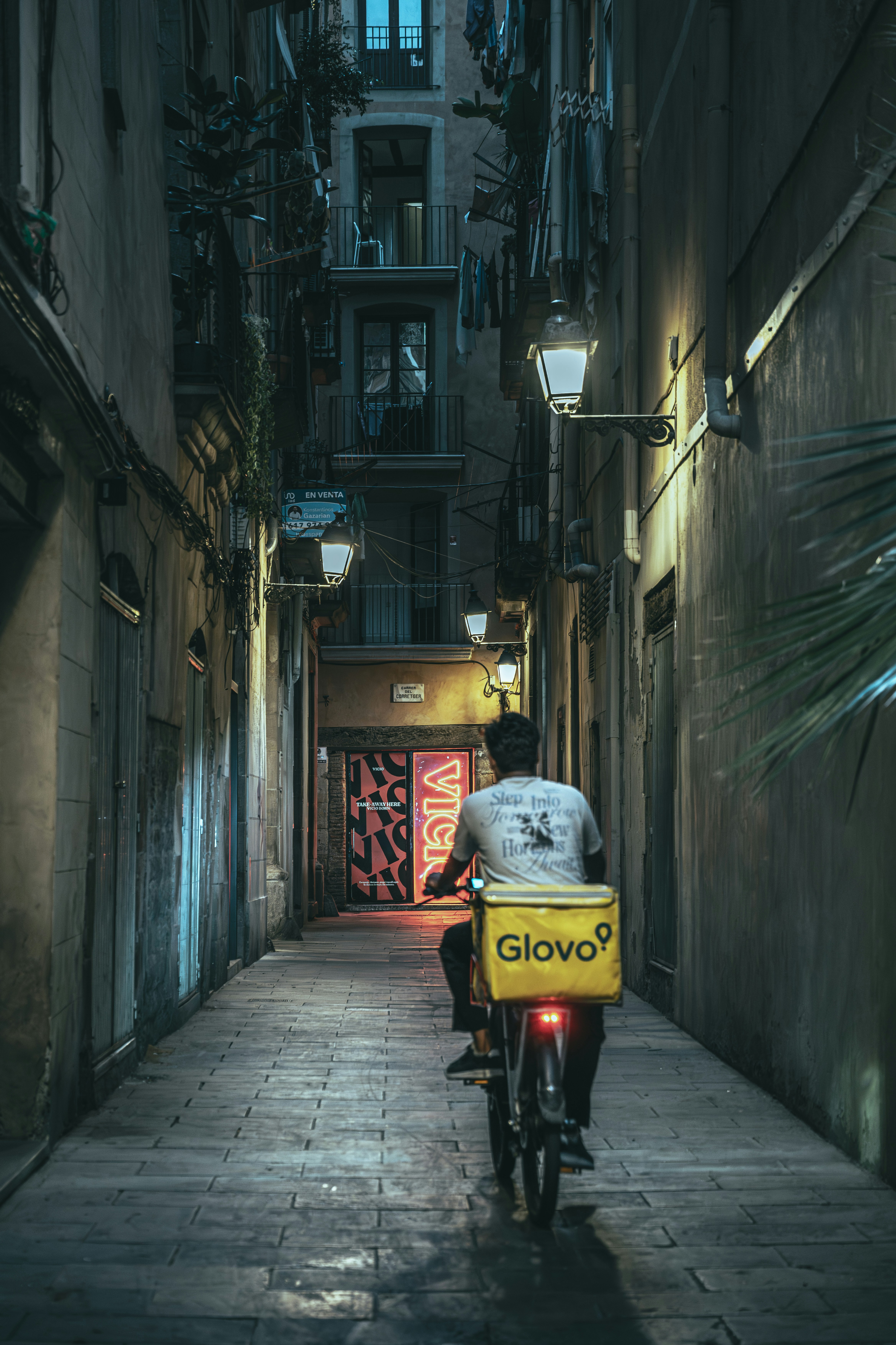 Nighttime alley with a delivery rider on a yellow Glovo box traversing wet cobblestones toward a lit doorway; moody blue-green tones and urban textures define the scene.