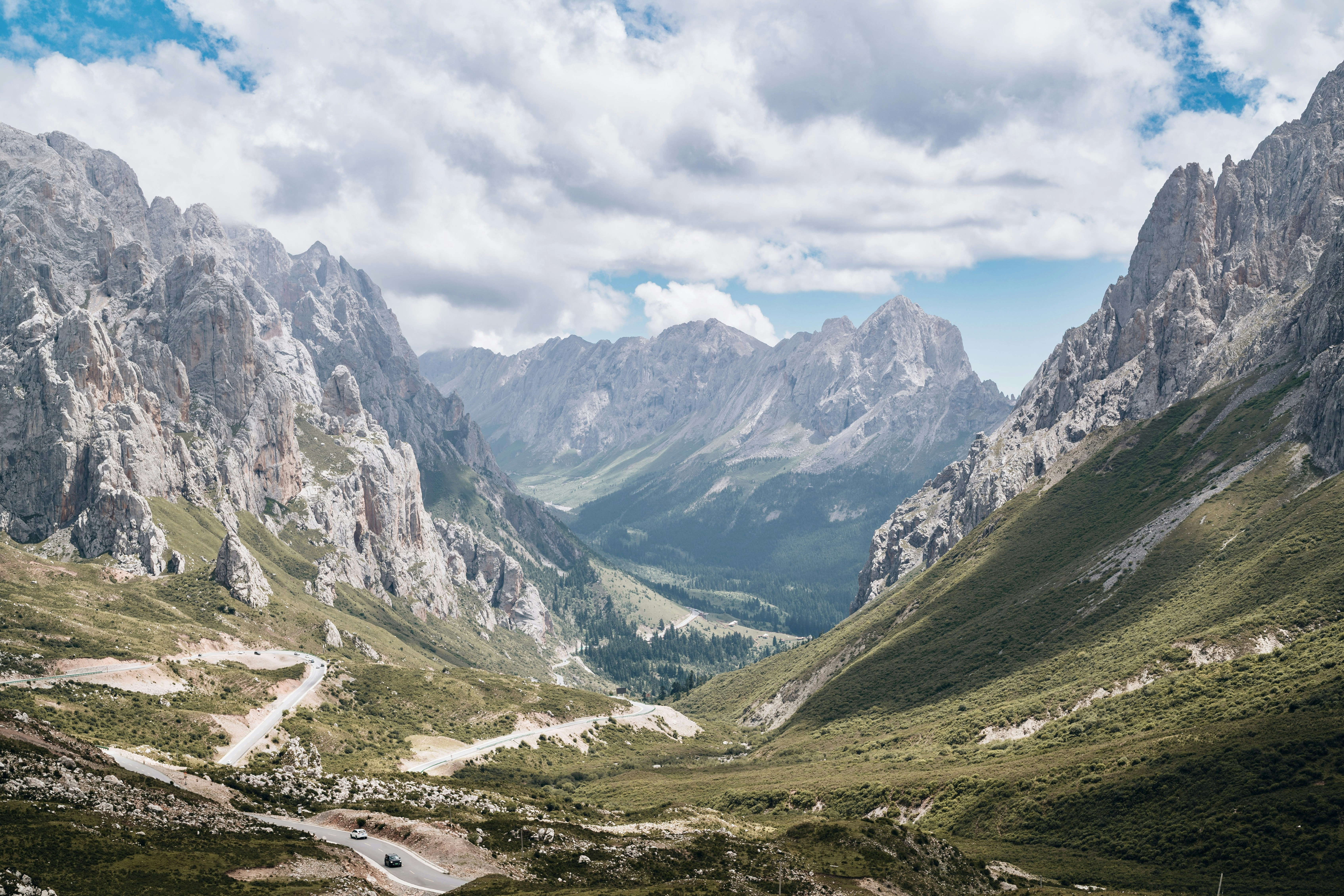 A panoramic valley carved by towering mountain peaks, with a winding dirt road cutting through grassy slopes toward a distant valley under a partly cloudy sky.