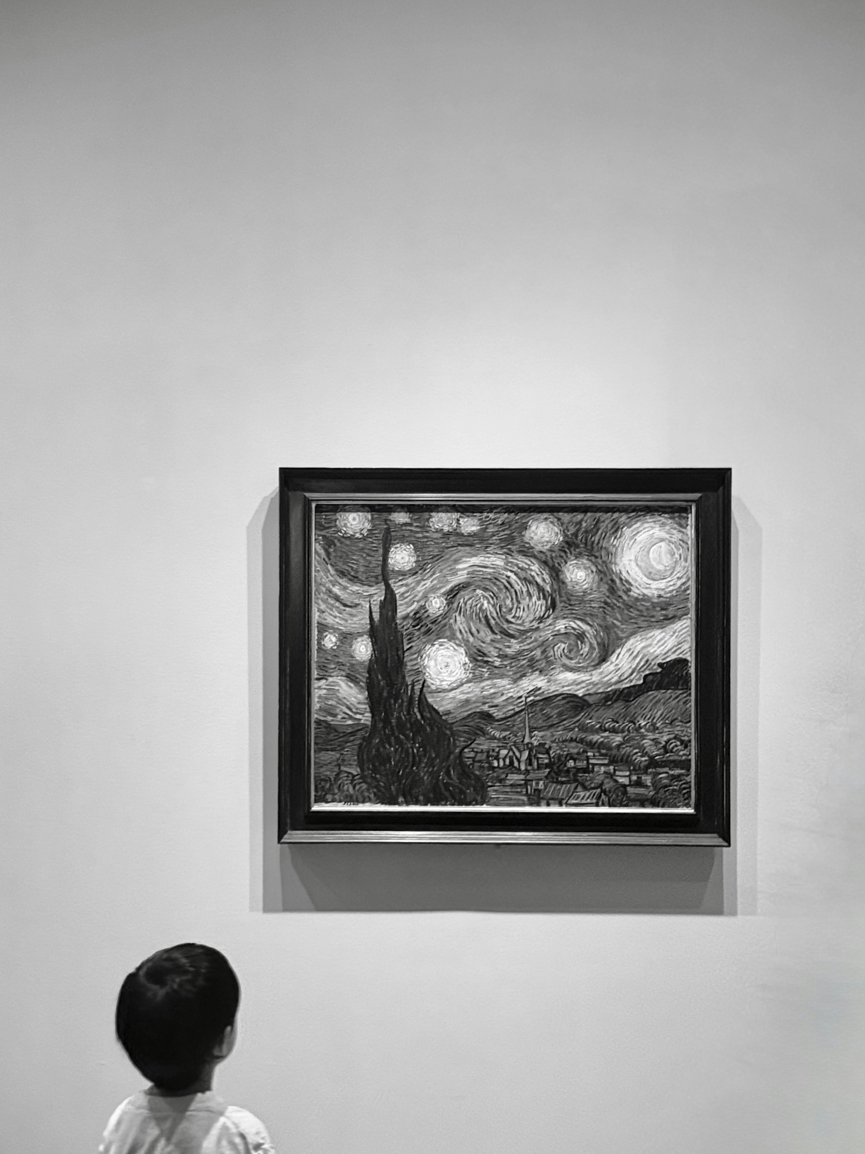 A young child stands in front of a framed Vincent van Gogh–style painting on a gallery wall, observing the swirling night sky imagery. The piece is mounted with a dark frame in a white-walled space.