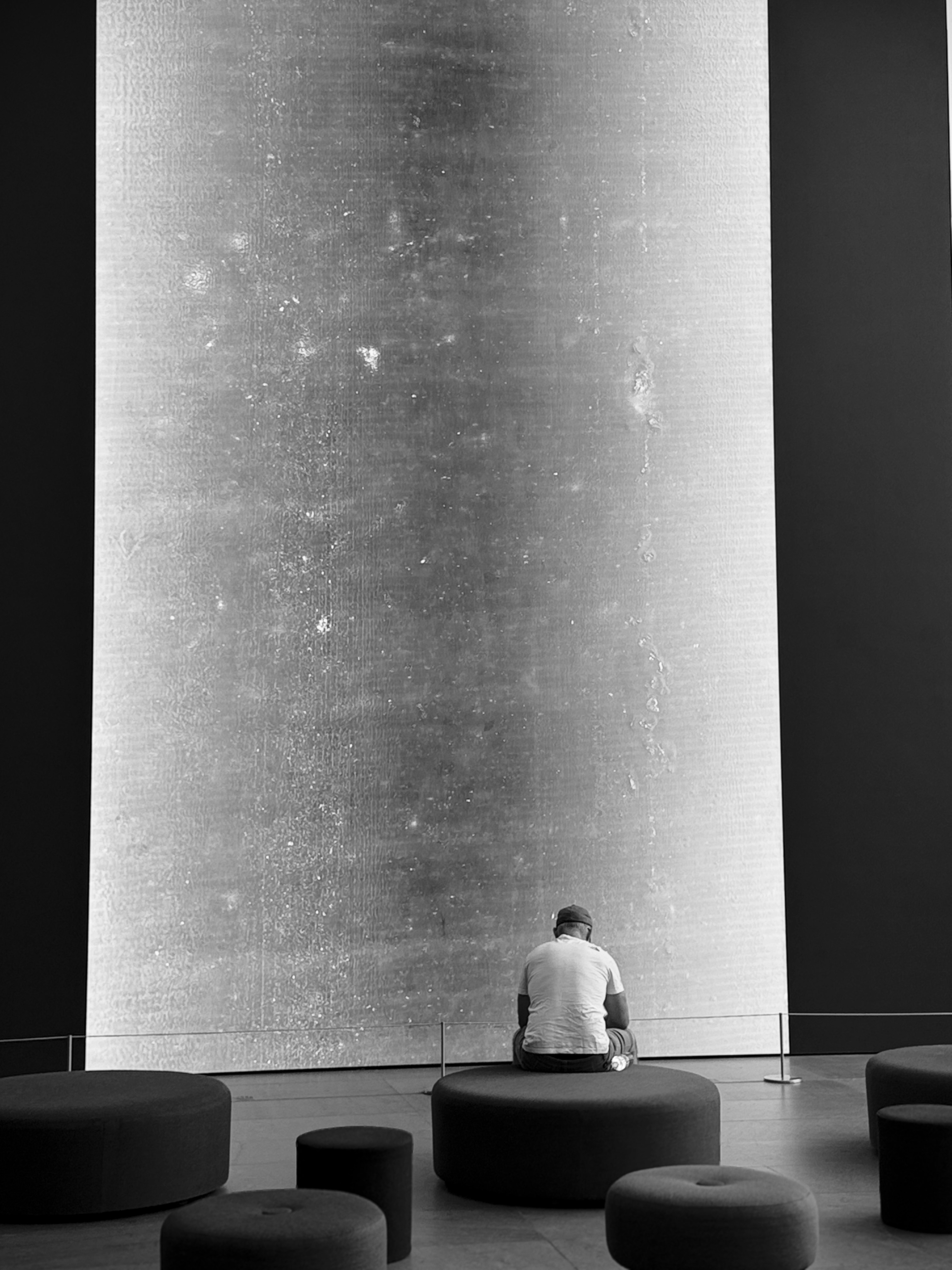 A person in a t-shirt sitting on a circular stool before a large screen