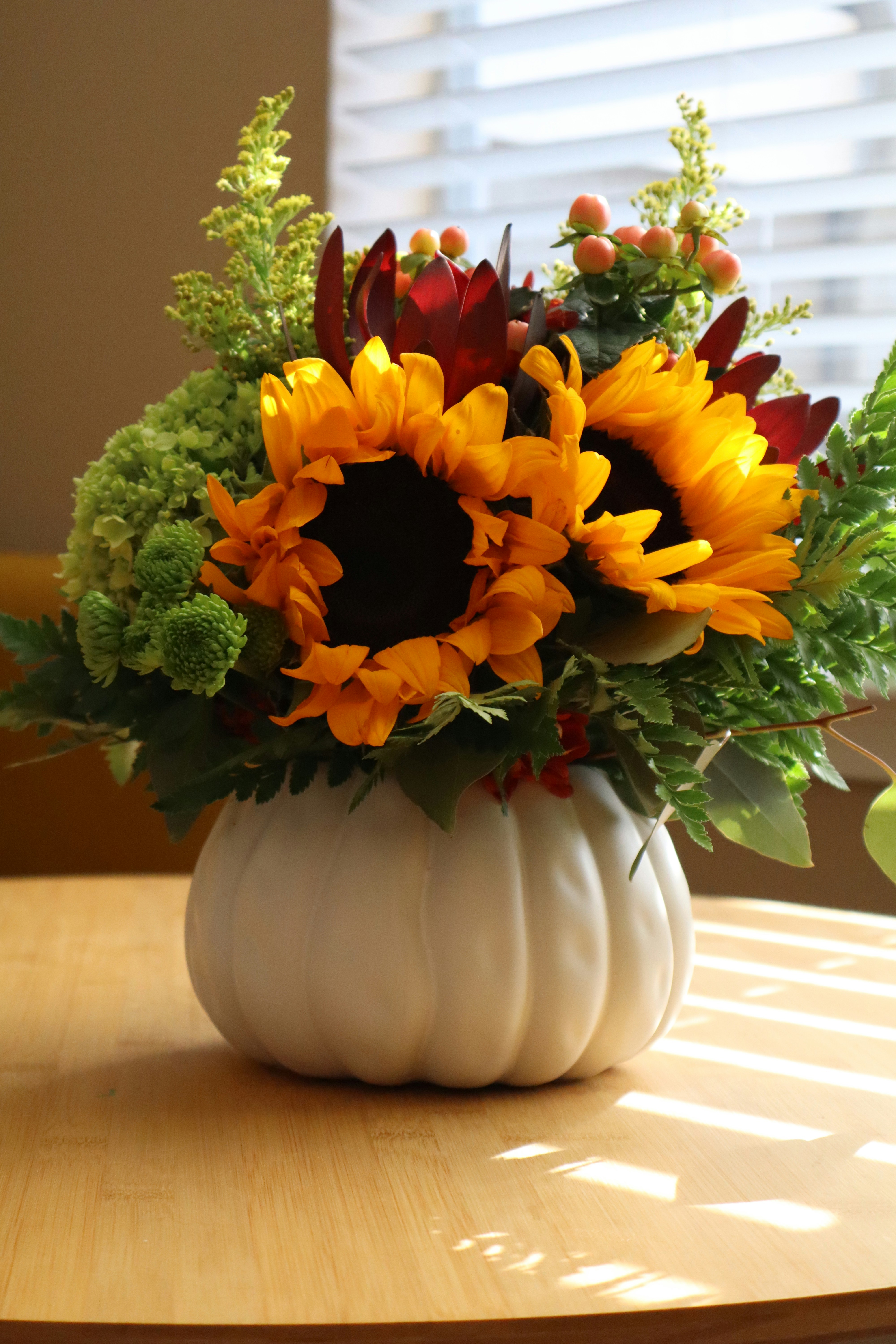 Bright bouquet featuring sunflowers in a white pumpkin vase bathed in warm window light.