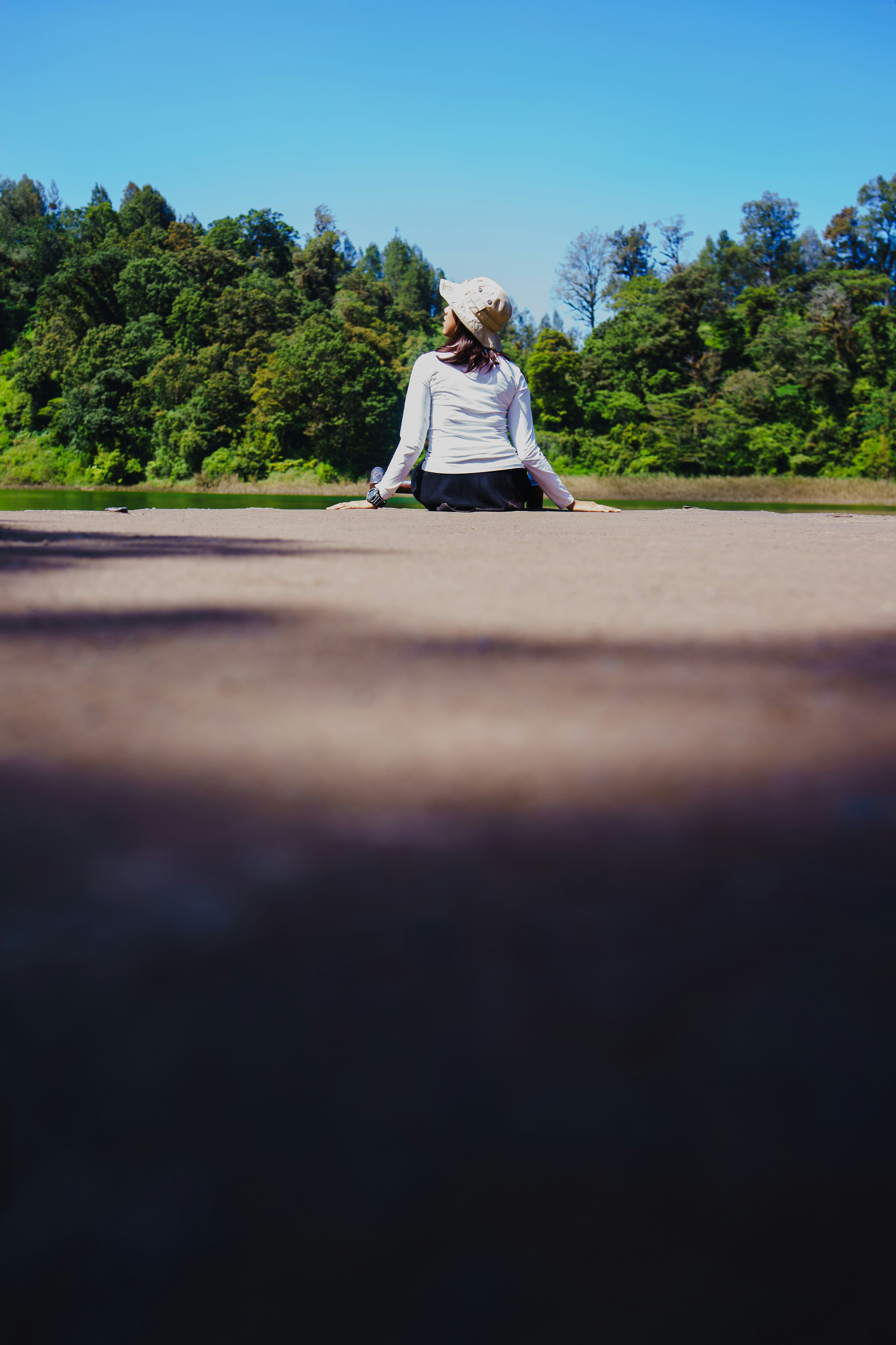 A person sits cross-legged on a dock facing a calm lake with a lush tree line under a clear blue sky. This is a candid shot capturing quiet reflection; likely a lifestyle scene with natural daylight.
