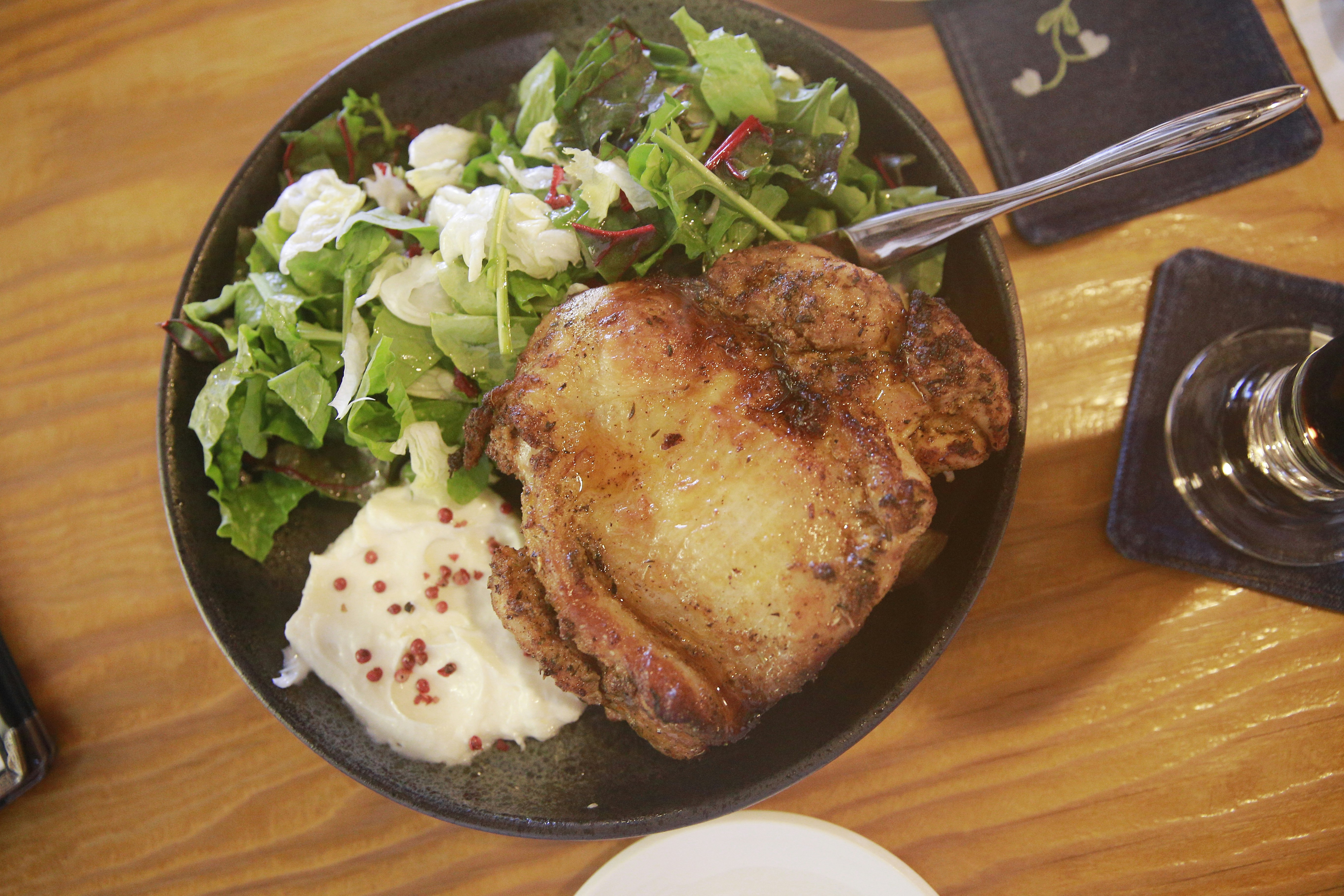Sautéed chicken thigh with a golden crust sits beside a fresh salad and a dollop of cream on a dark plate.